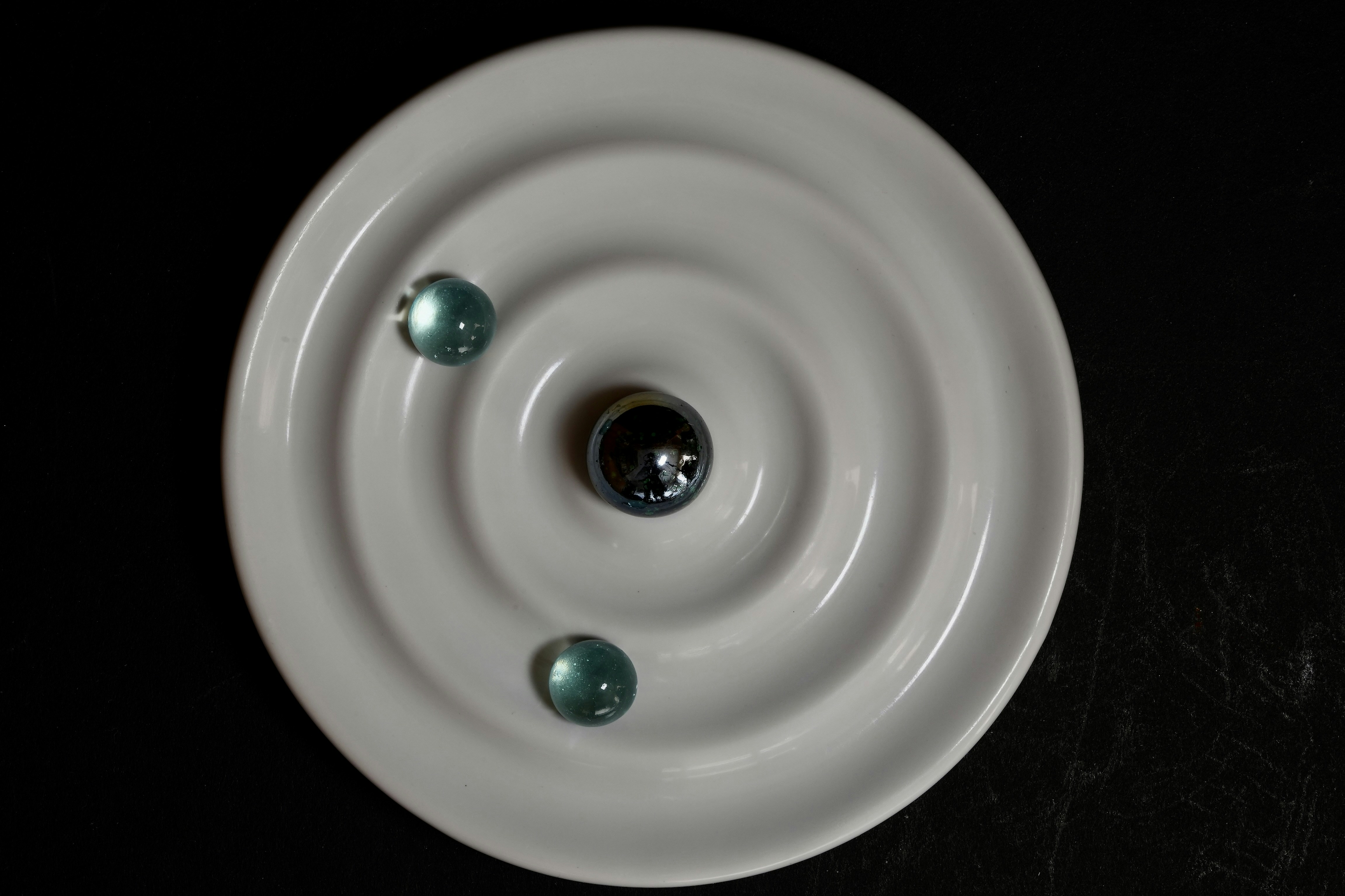 Close-up of a white ceramic plate with concentric circular depressions and two teal glass beads, set against a dark surface.