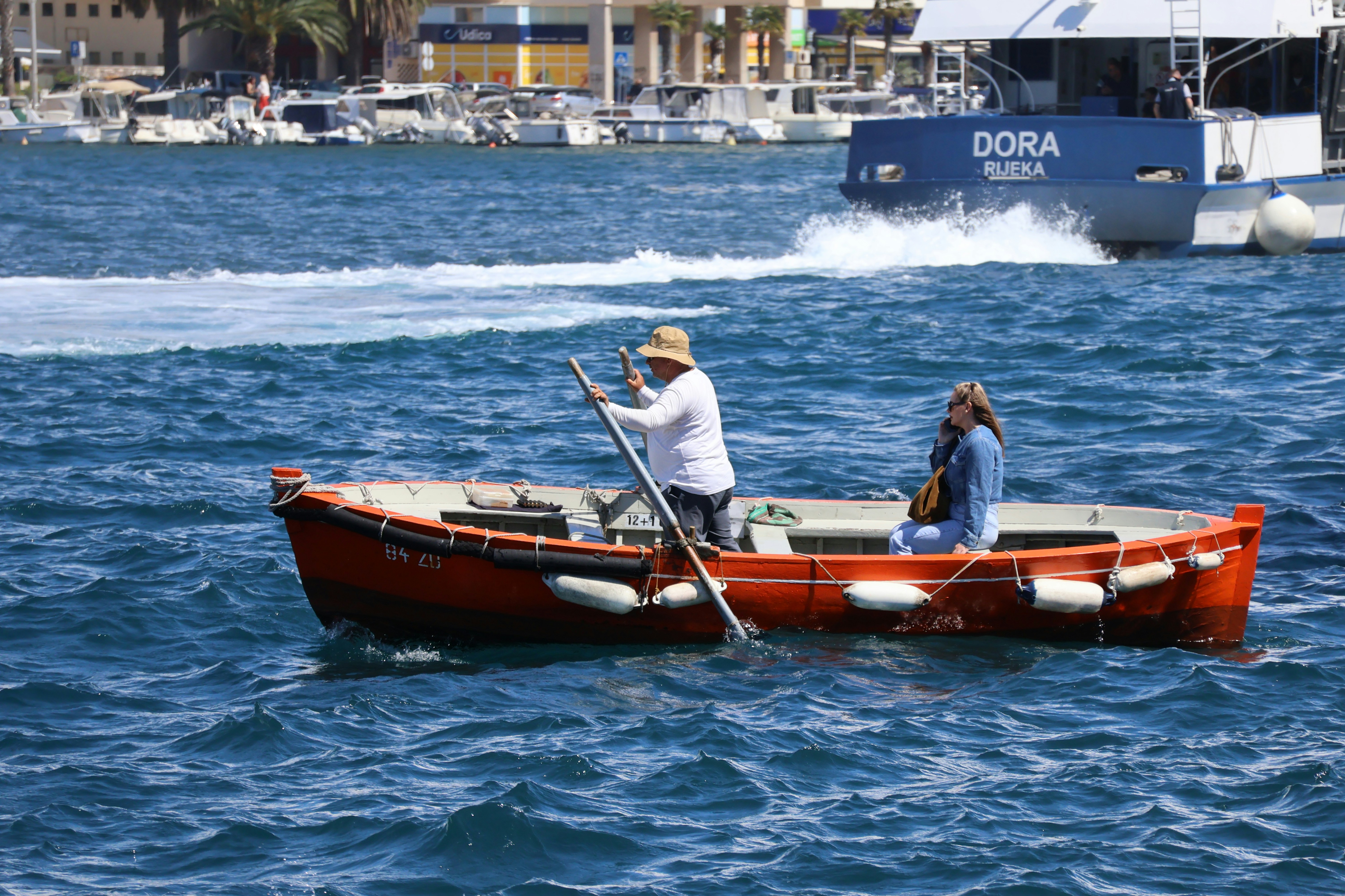 Two fishermen ride a small orange boat across sunlit harbor waters
