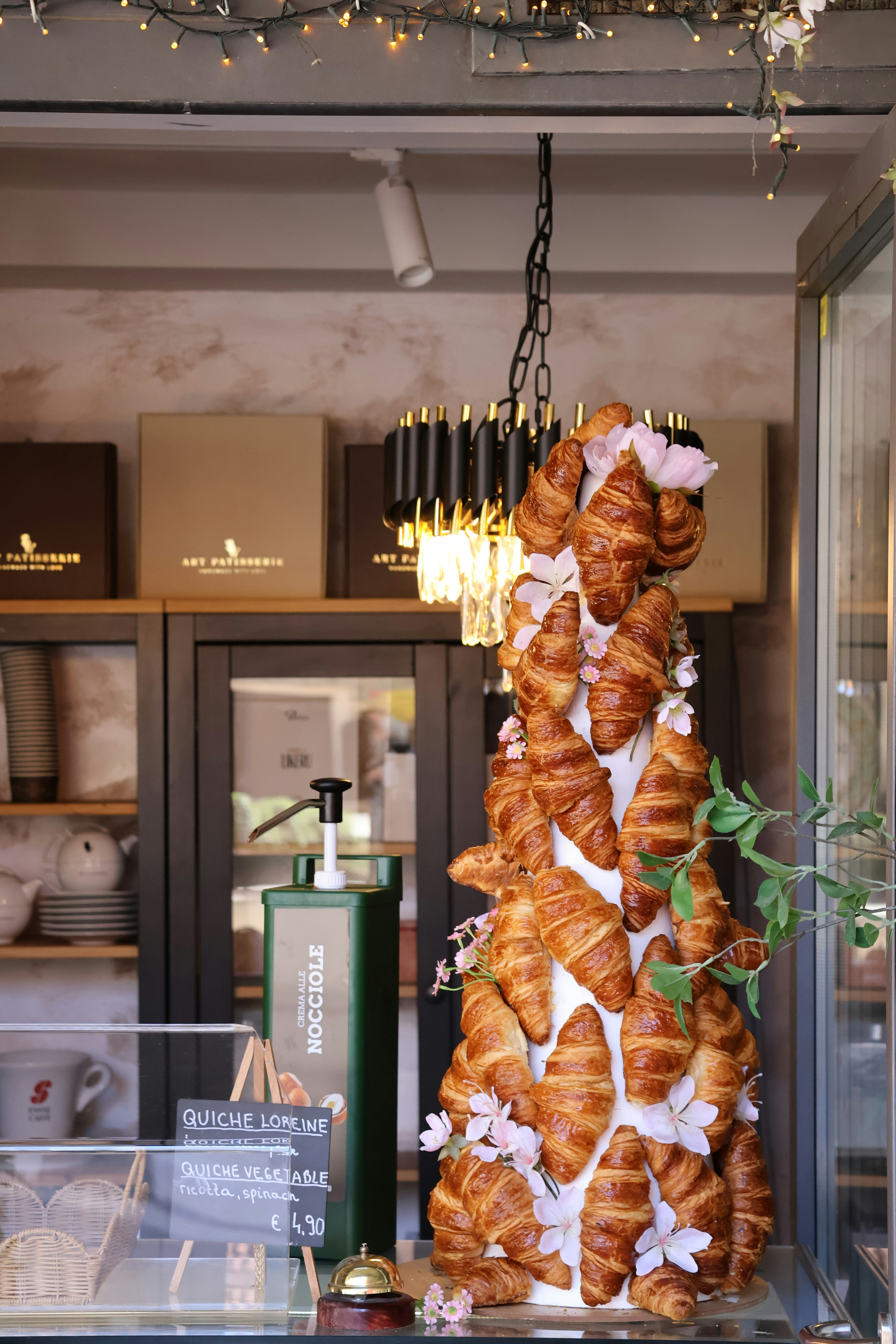 A tall croissant sculpture stacked in a shop window, adorned with flowers, against shelves of pastry boxes and a green soap dispenser; a storefront display emphasizing baked goods.