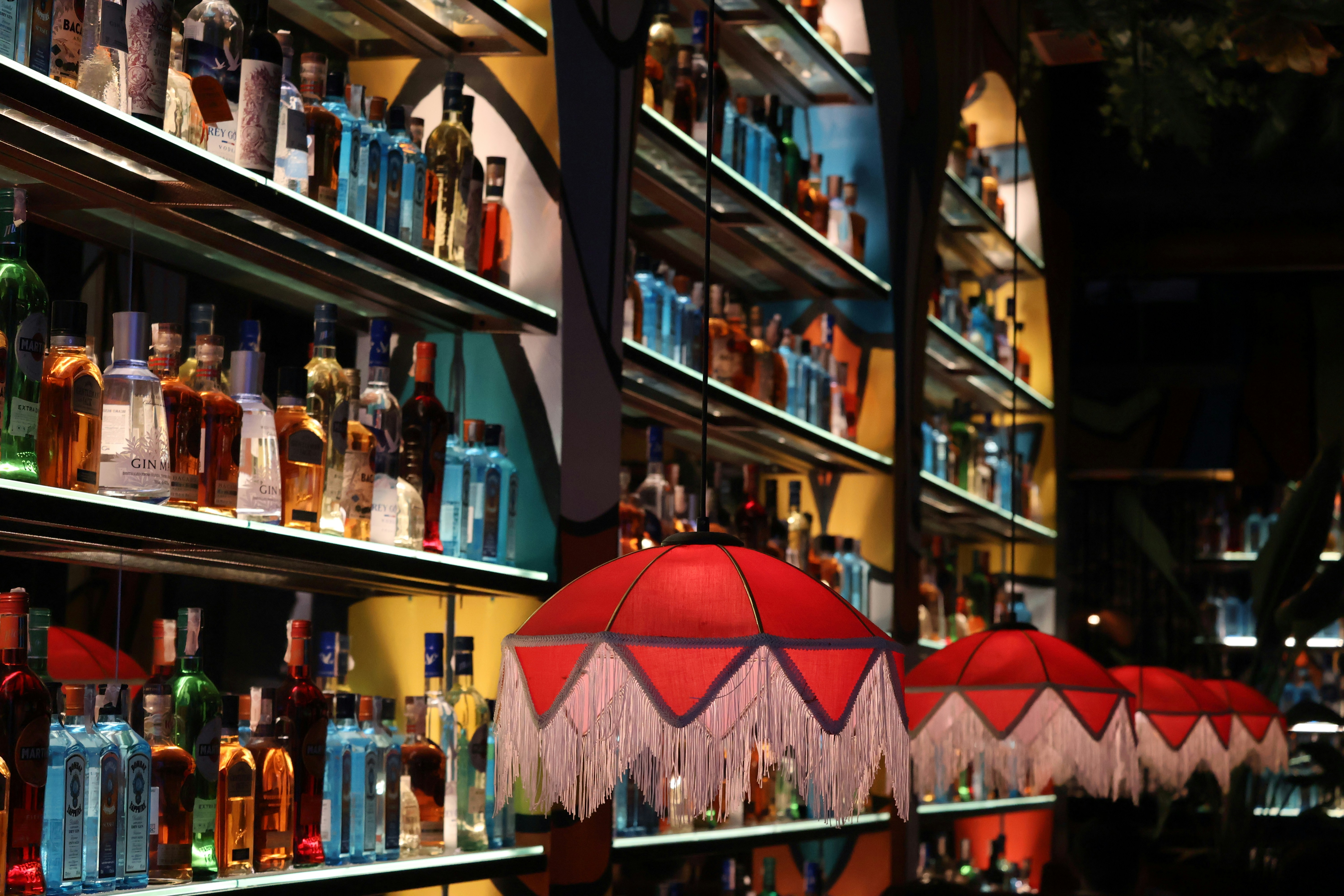 Vibrant liquor shelves glow along a colorful bar, with red parasol fringes adding a whimsical touch to a dim, retail-lit scene.