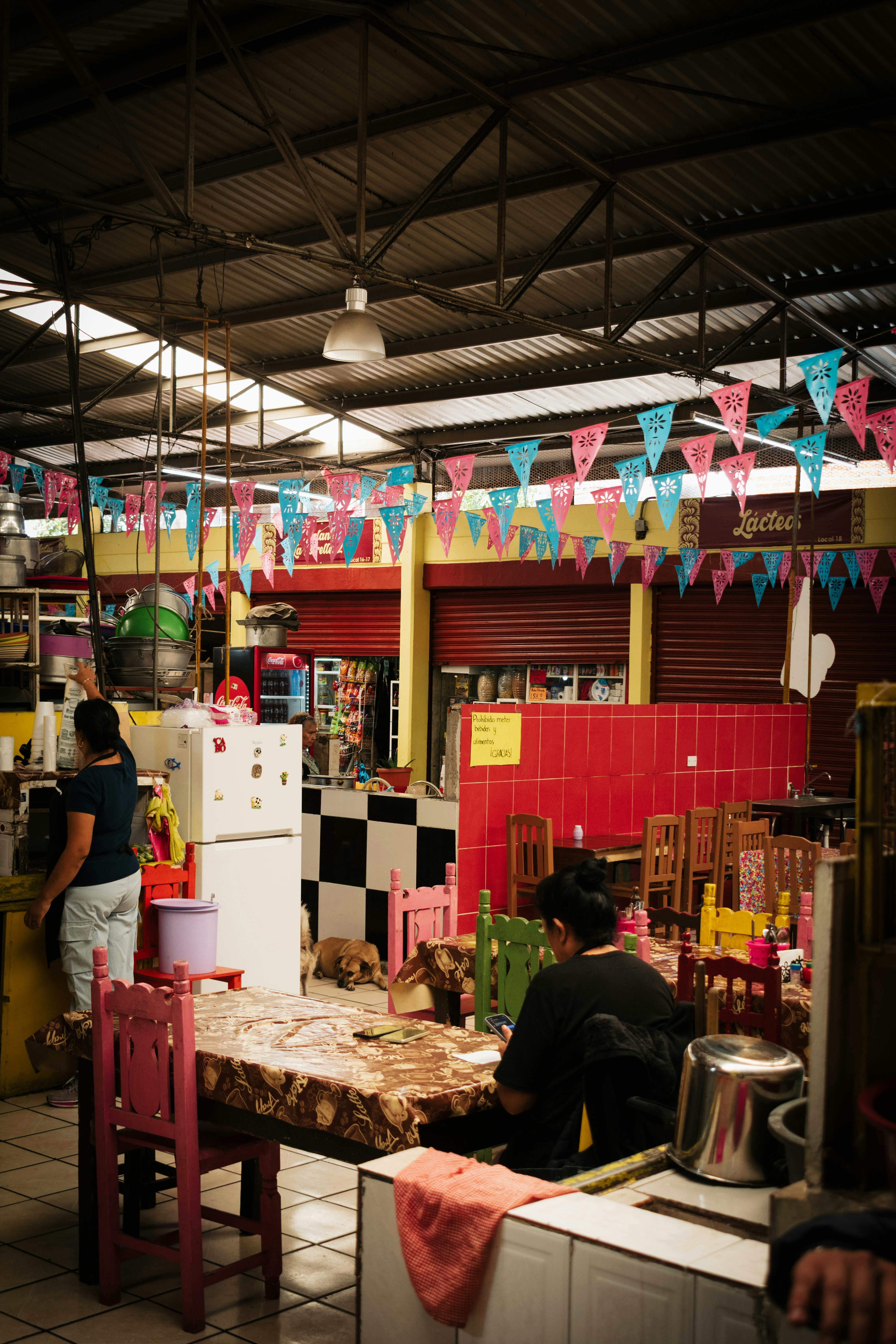 Vibrant market interior with colorful pennant banners, a tiled counter area, and diners at tables under a corrugated roof.