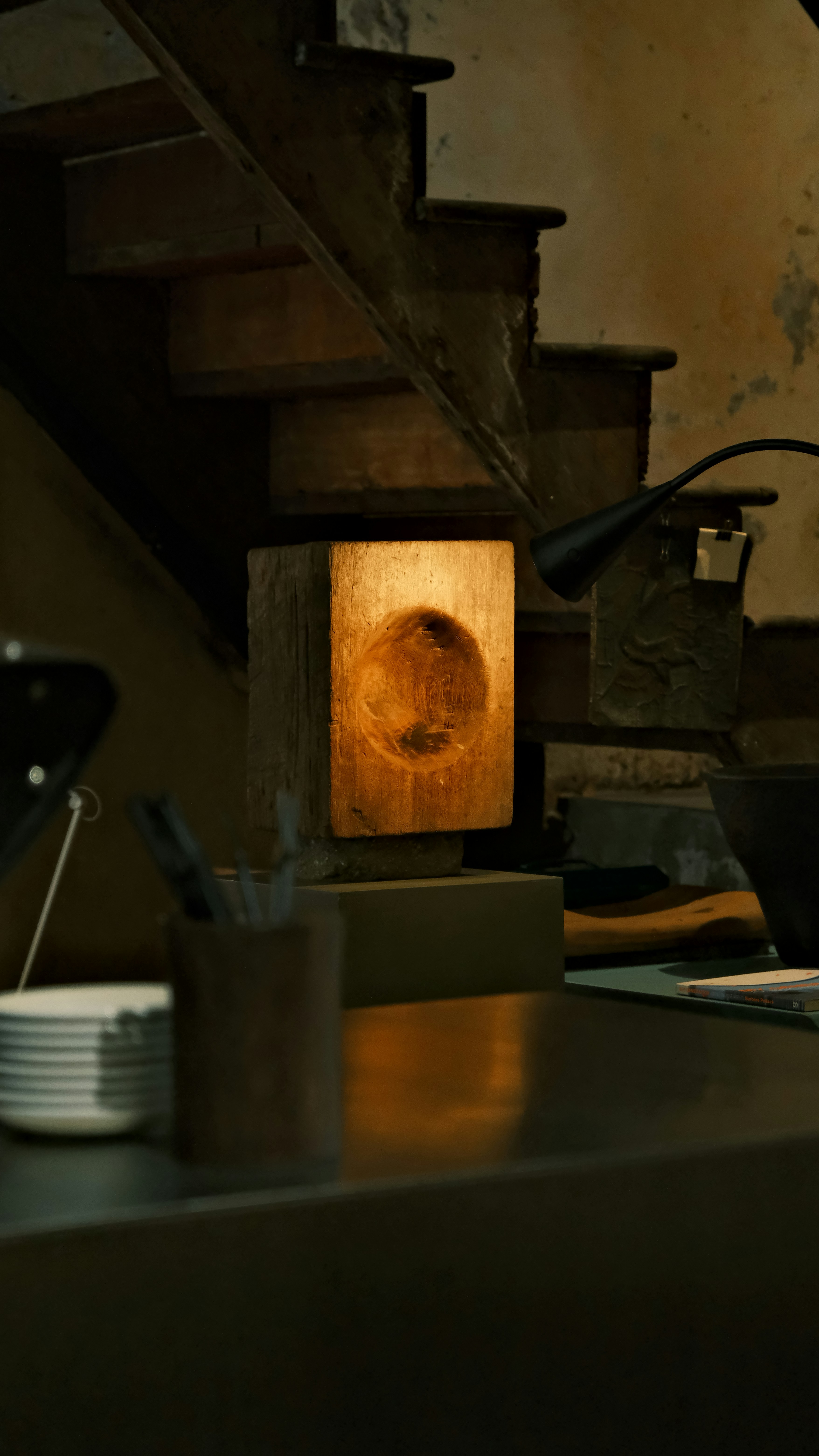 A close-up in a dim workshop shows a illuminated wooden block with a knot perched on a metal surface; surrounding tools and stairs recede into darkness, giving a moody, intimate glimpse of a crafting space. (photo is a still life/industrial interior).