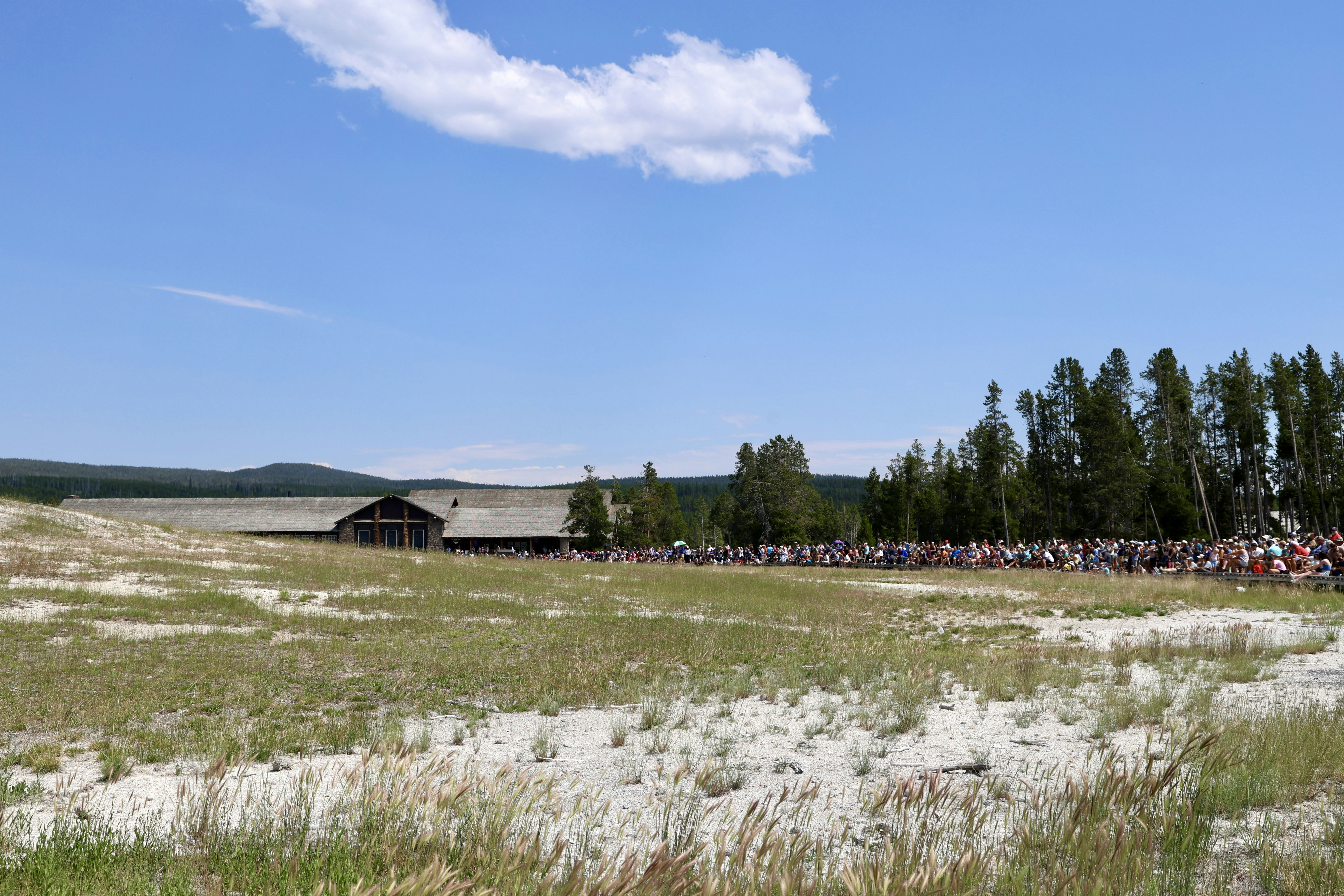 A broad field leads to a distant crowd gathered along a low structure, with a bright blue sky and scattered clouds above.