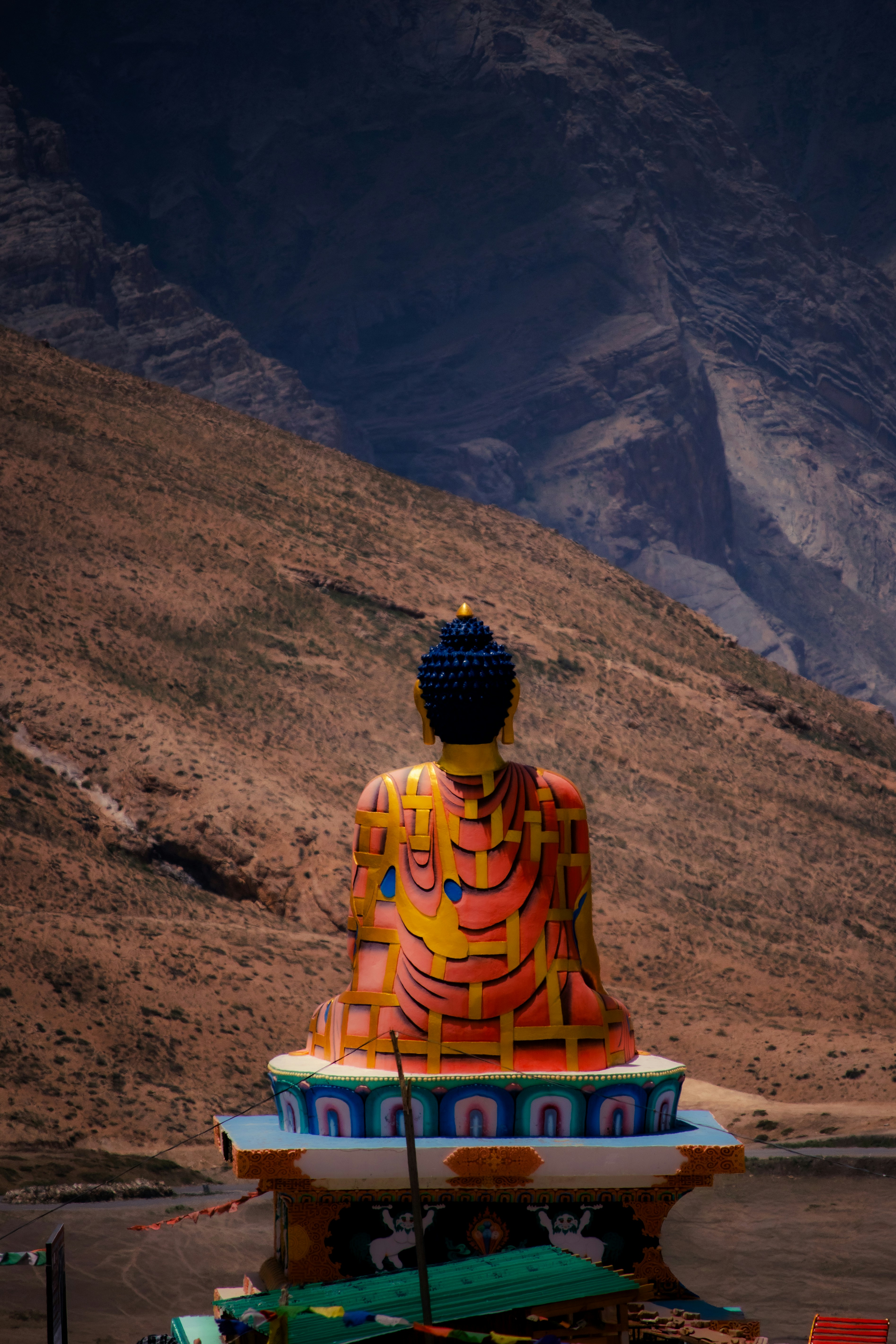 Vibrant Buddha statue seated on a painted pedestal in a barren valley with rugged hills in the background. The scene emphasizes color and sculpture against a stark, earthy landscape.