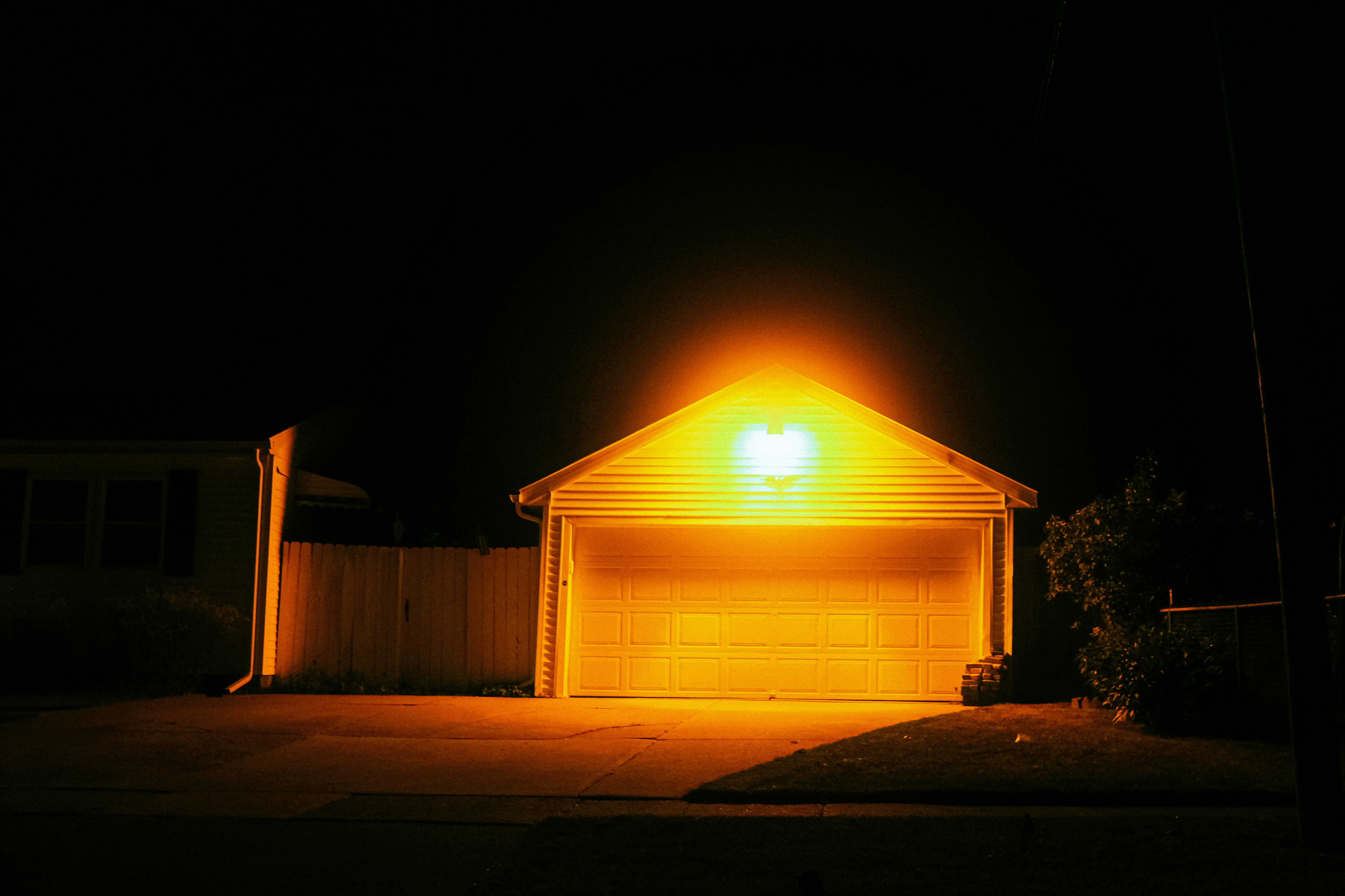 A dimly lit suburban garage at night bathed in amber light from a bright fixture above the door. The scene emphasizes the house front and driveway with soft shadows and a calm, nocturnal mood.