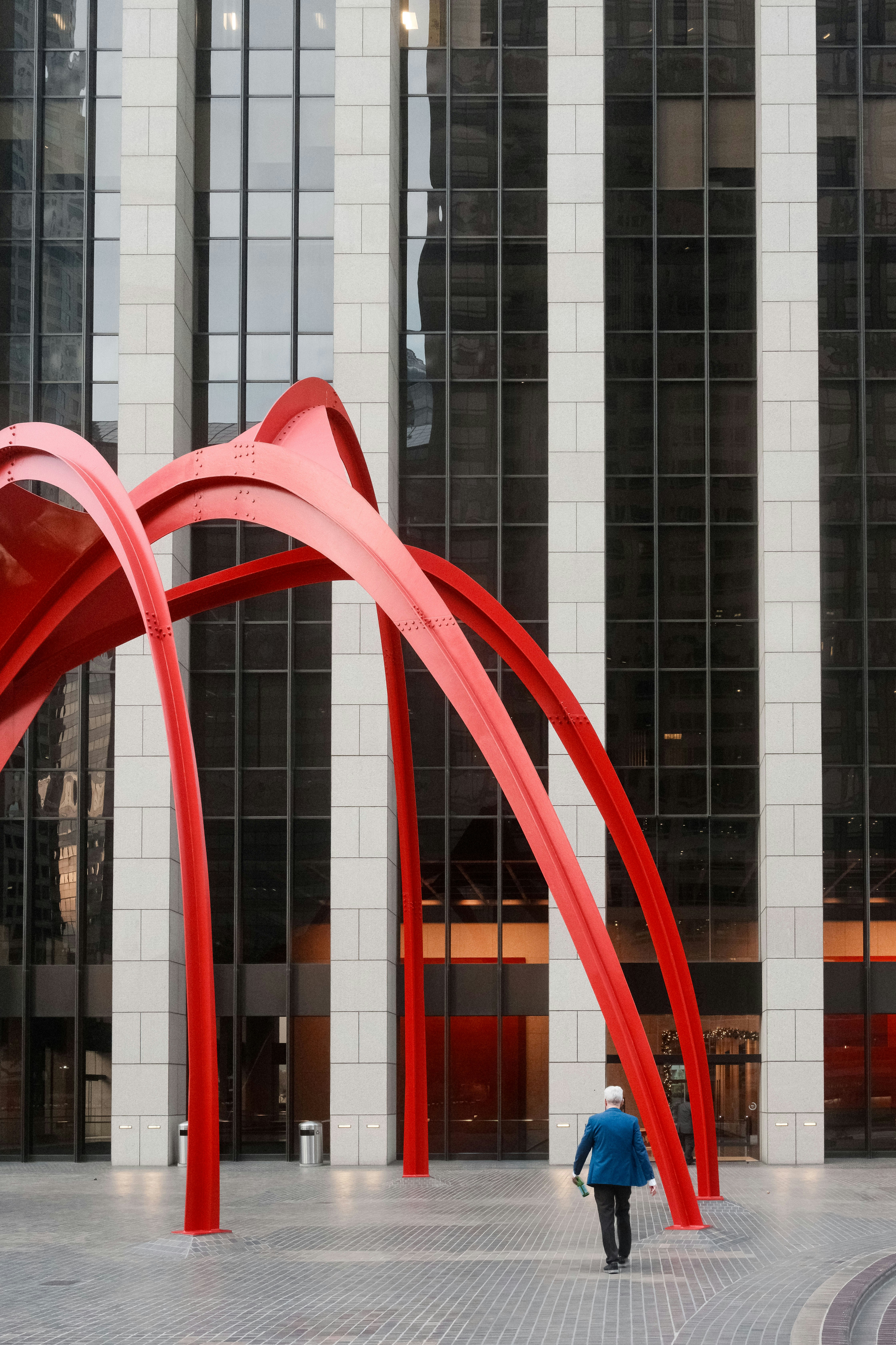 Red sculpture arches rise beside a glass-fronted building, with a lone person in a blue jacket strolling nearby.