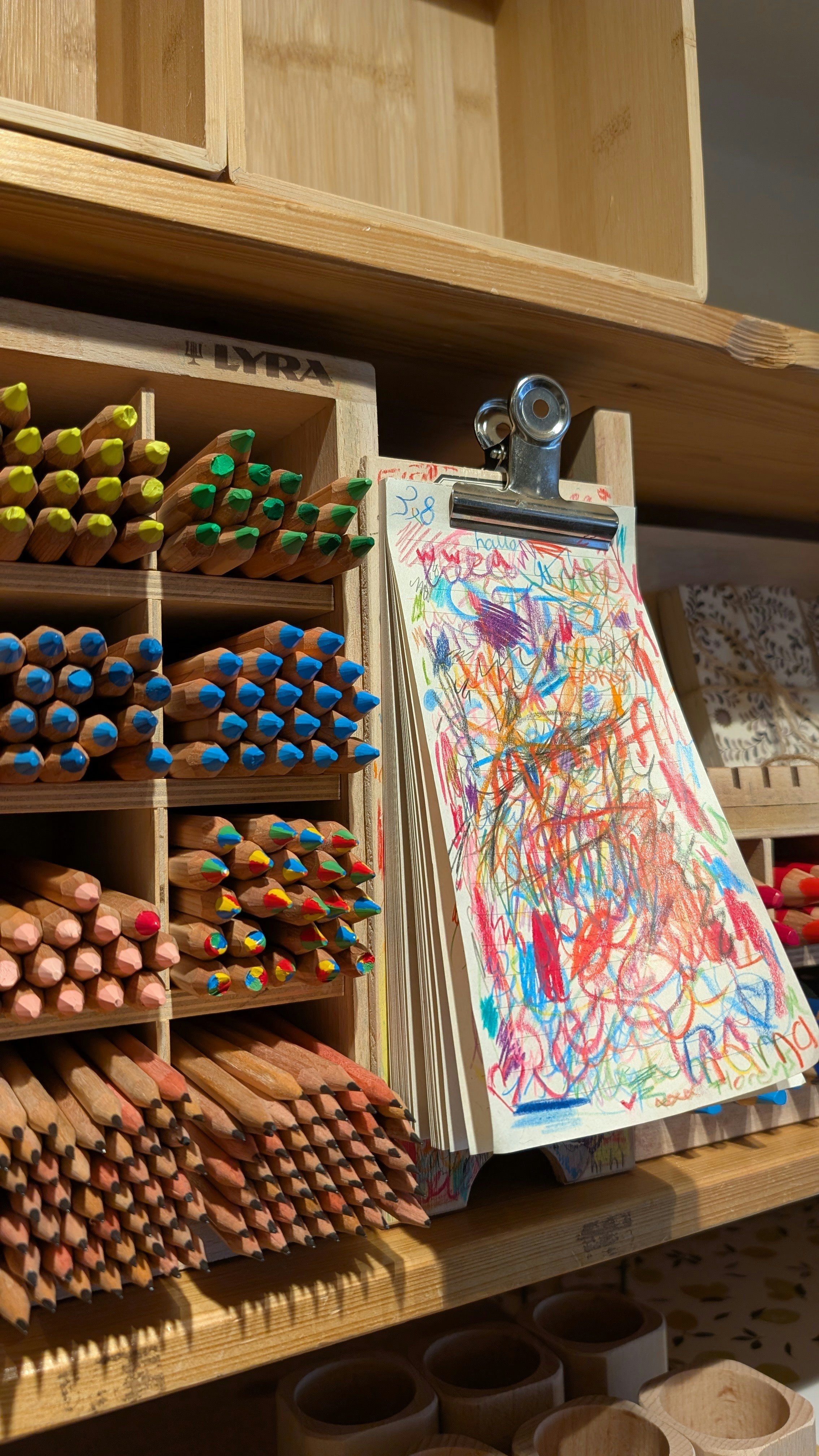Colorful pencils organized in wooden cubbies frame a scribbled drawing on a clipboard, capturing a cozy art corner ready for creativity.