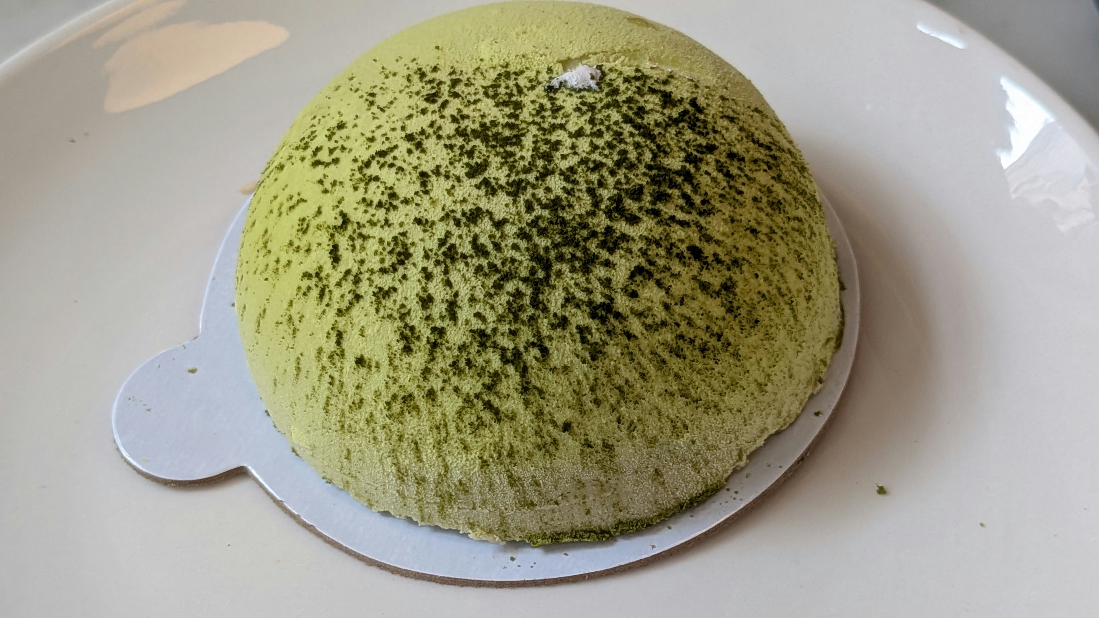 Green matcha mousse cake sits on a white plate with a decorative tab, photographed from a slightly downward angle to emphasize texture.