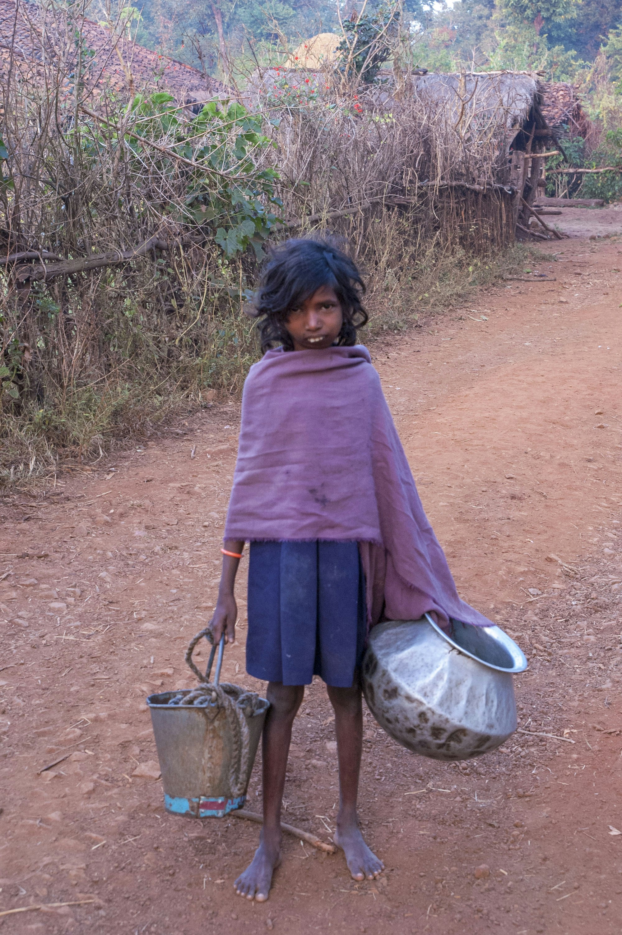 A young girl stands barefoot on a dusty road, carrying a metal pot and a smaller bucket, with a purple shawl draped over her shoulders. The rural village setting shows thatched huts and dry vegetation in the background.