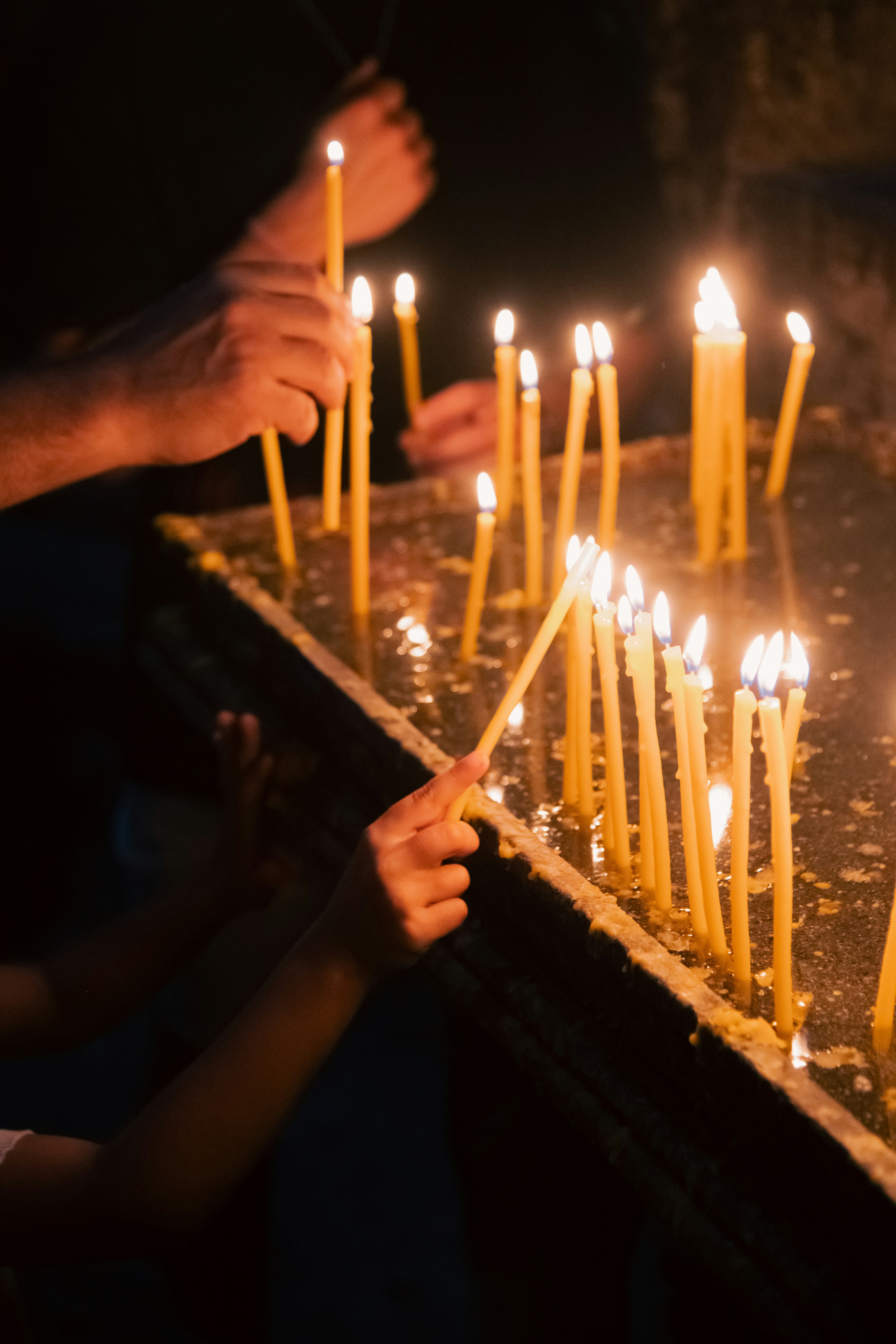 A group of hands lighting slender candles along a dim stone ledge, glow from the flames casting warm, intimate light. Photograph captures a communal moment at a ritual, with soft bokeh in the background.
