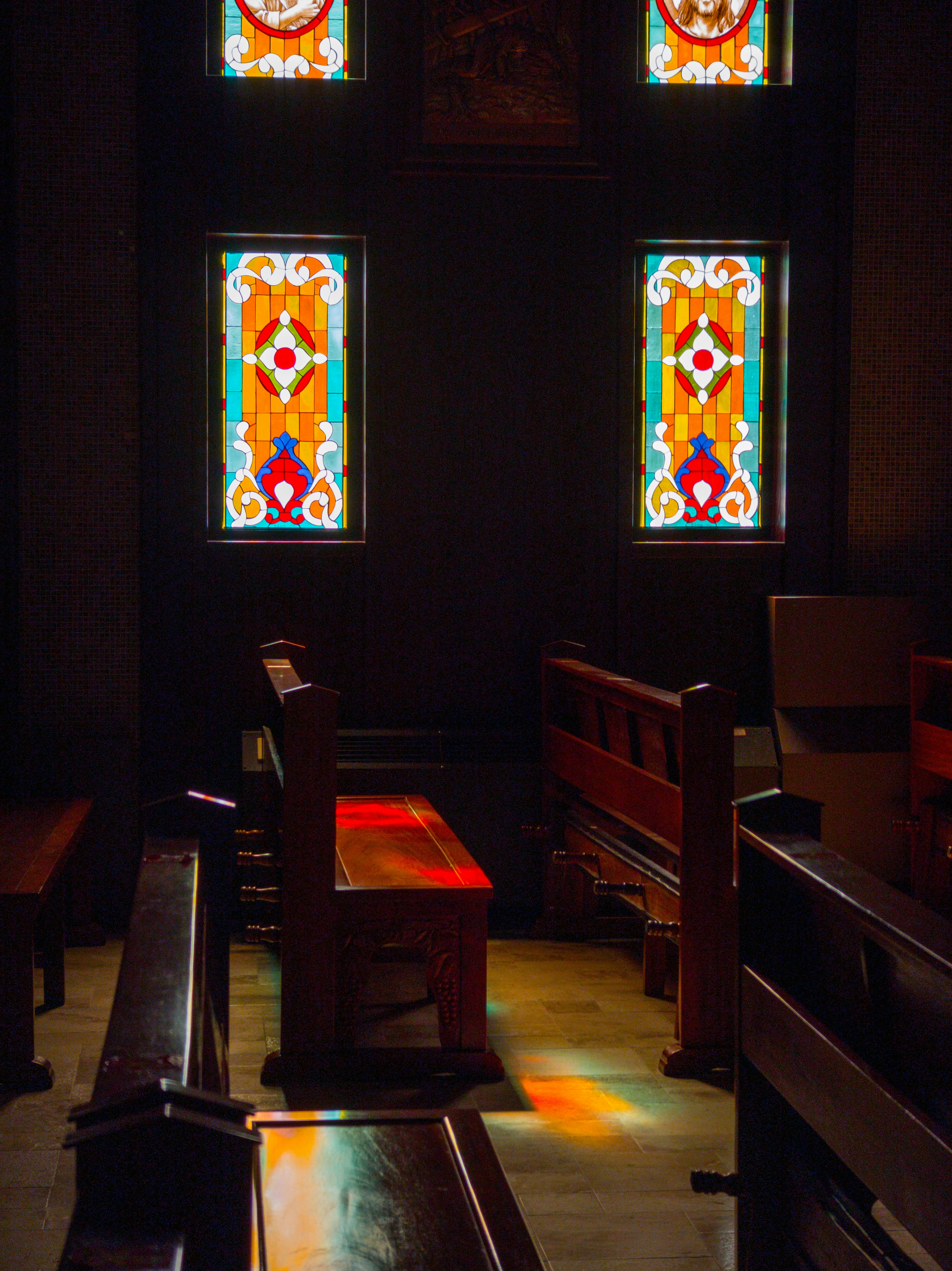 Dark interior of a small chapel with illuminated stained-glass panels casting colorful reflections on wooden pews and floor.