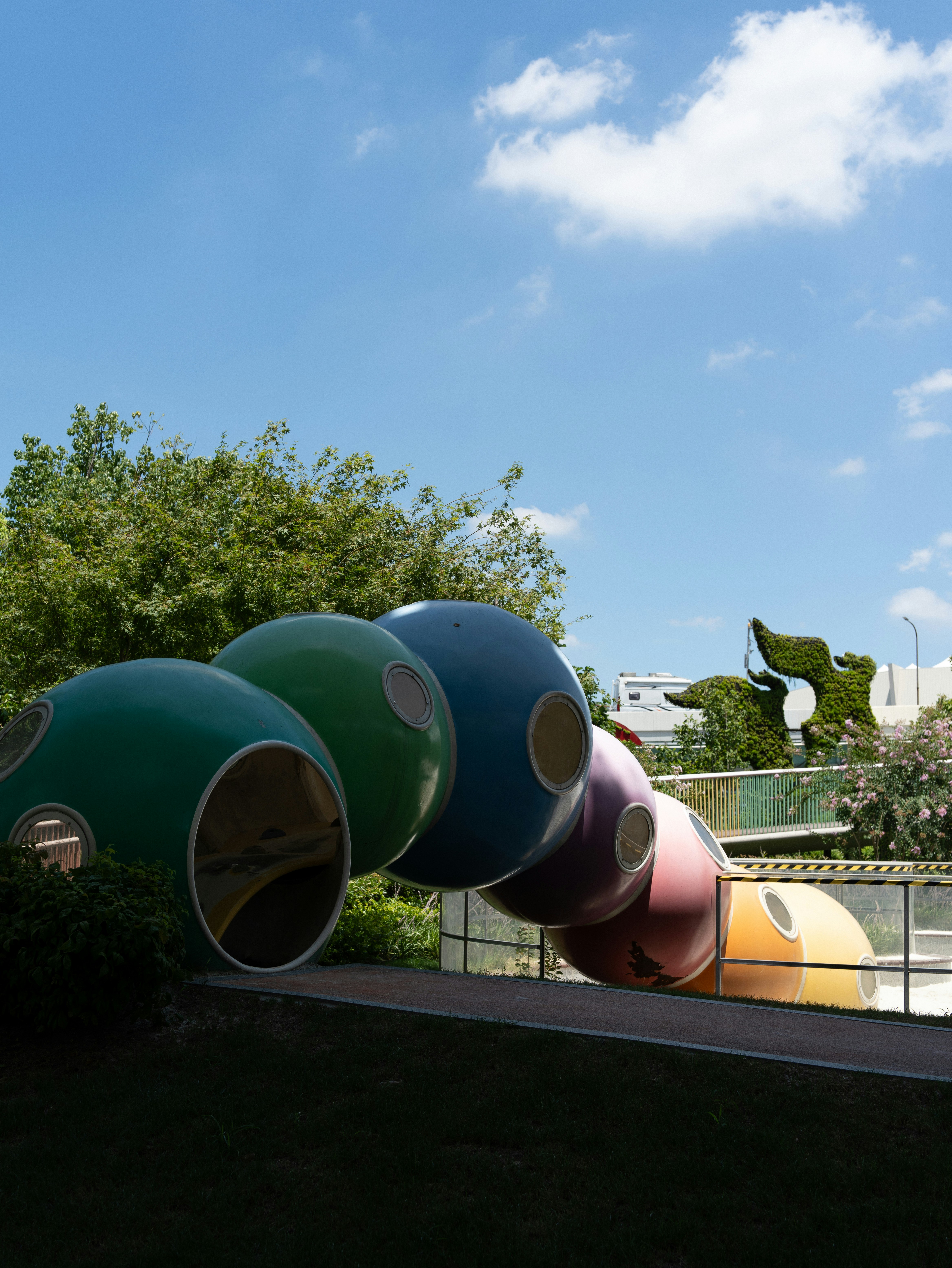 Colorful oversized tubes form a whimsical playground sculpture under a clear blue sky, with surrounding greenery and a distant sculpted hedge in view.