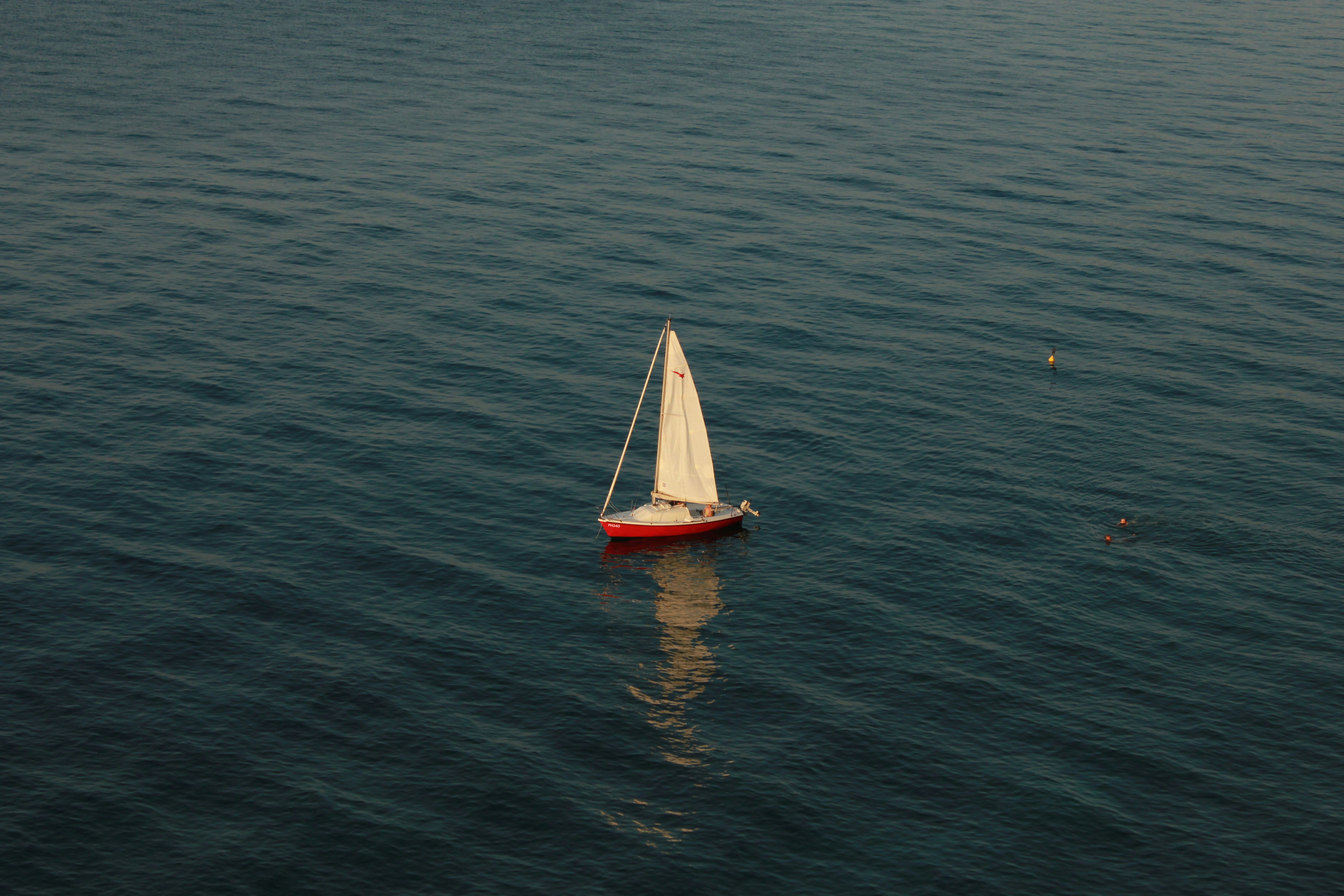 Red-hulled sailboat drifts on calm blue water with its white sail catching a light breeze; small waves and a lone buoy add context to a tranquil seascape.