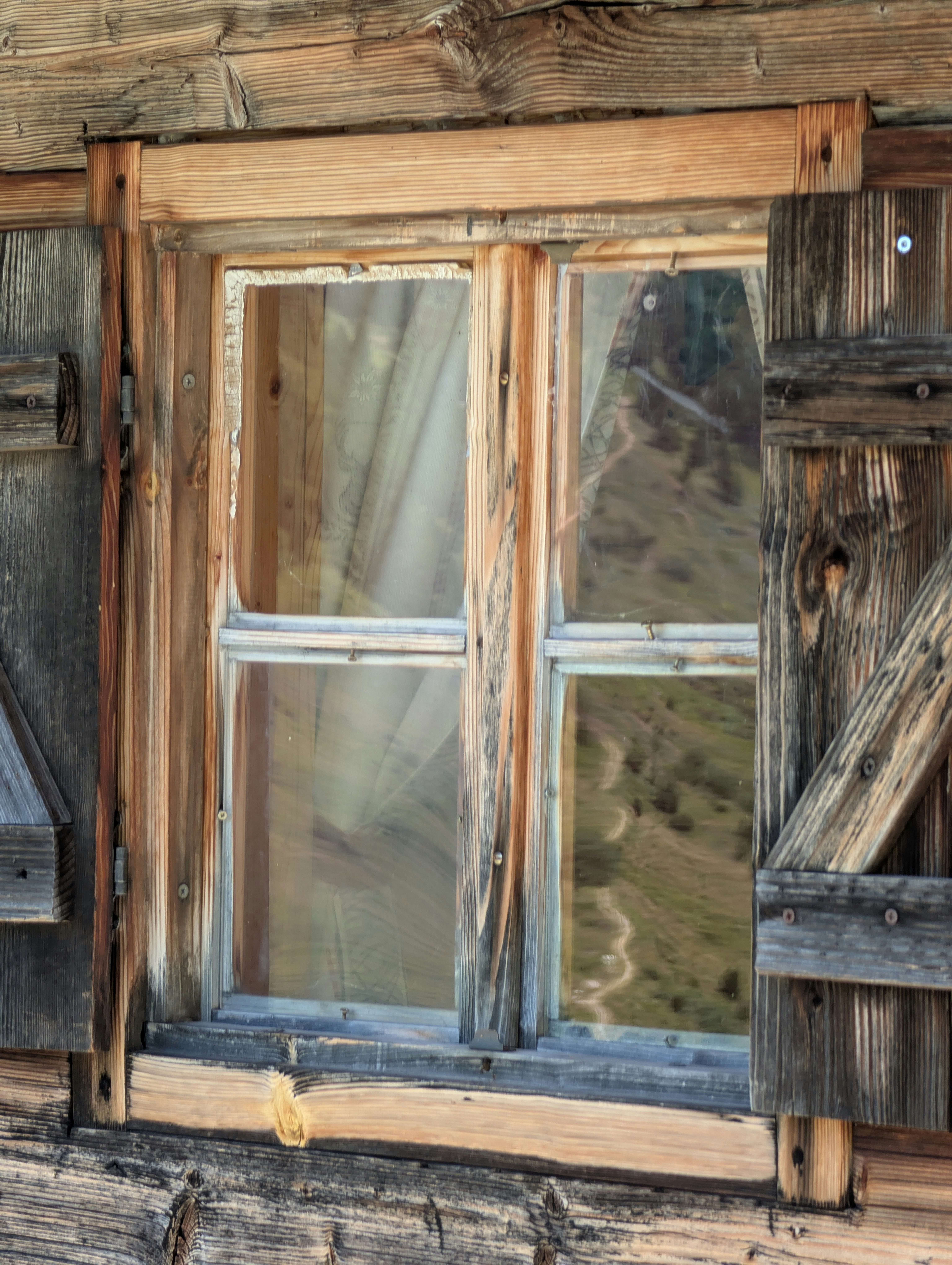 Weathered wooden window with peeling paint reveals a hint of a winding path outside, framed by aged shutters and rustic planks.