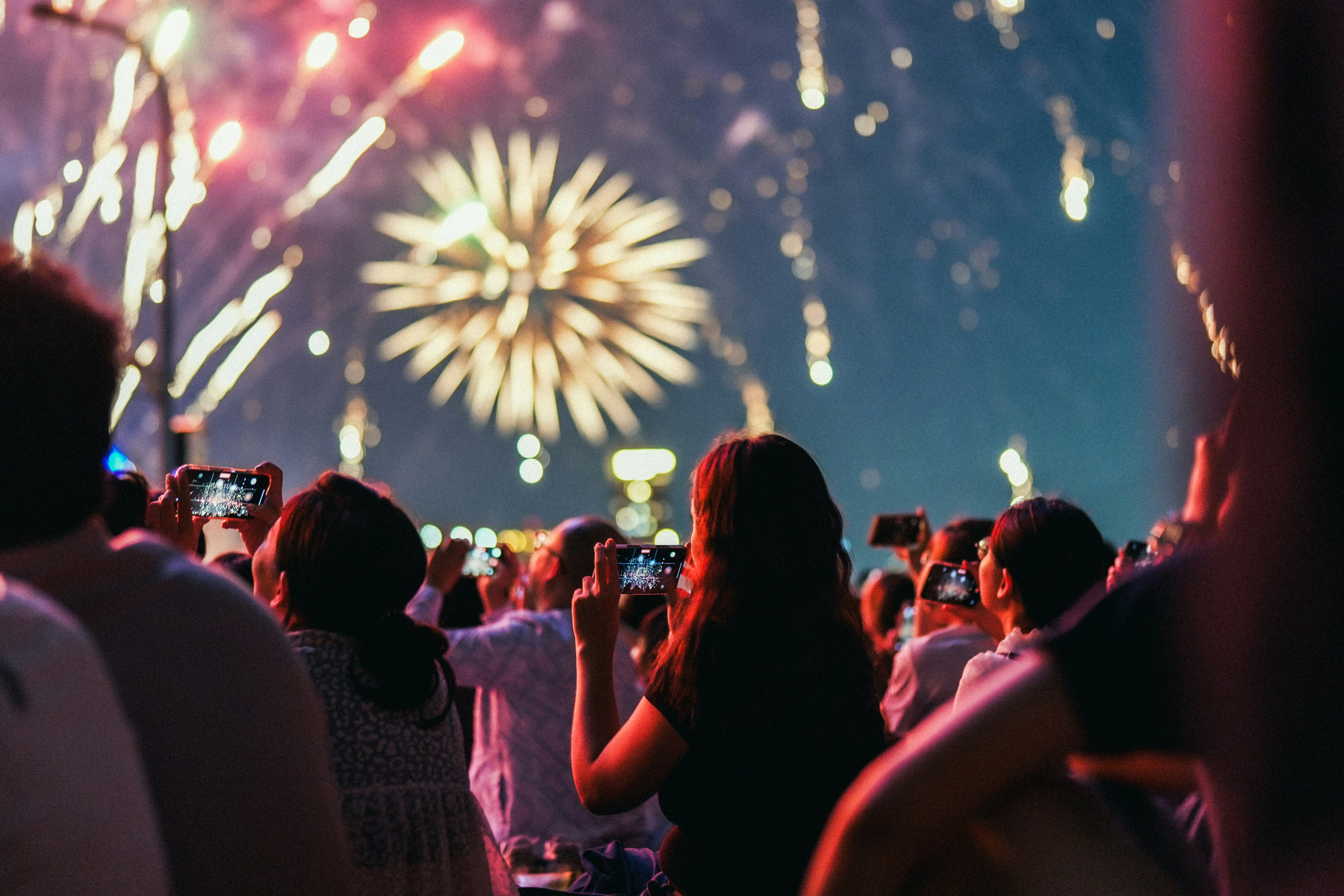 Crowd of spectators recording a fireworks display with smartphones as bursts illuminate the night sky; event scene emphasizing collective capture and celebration.