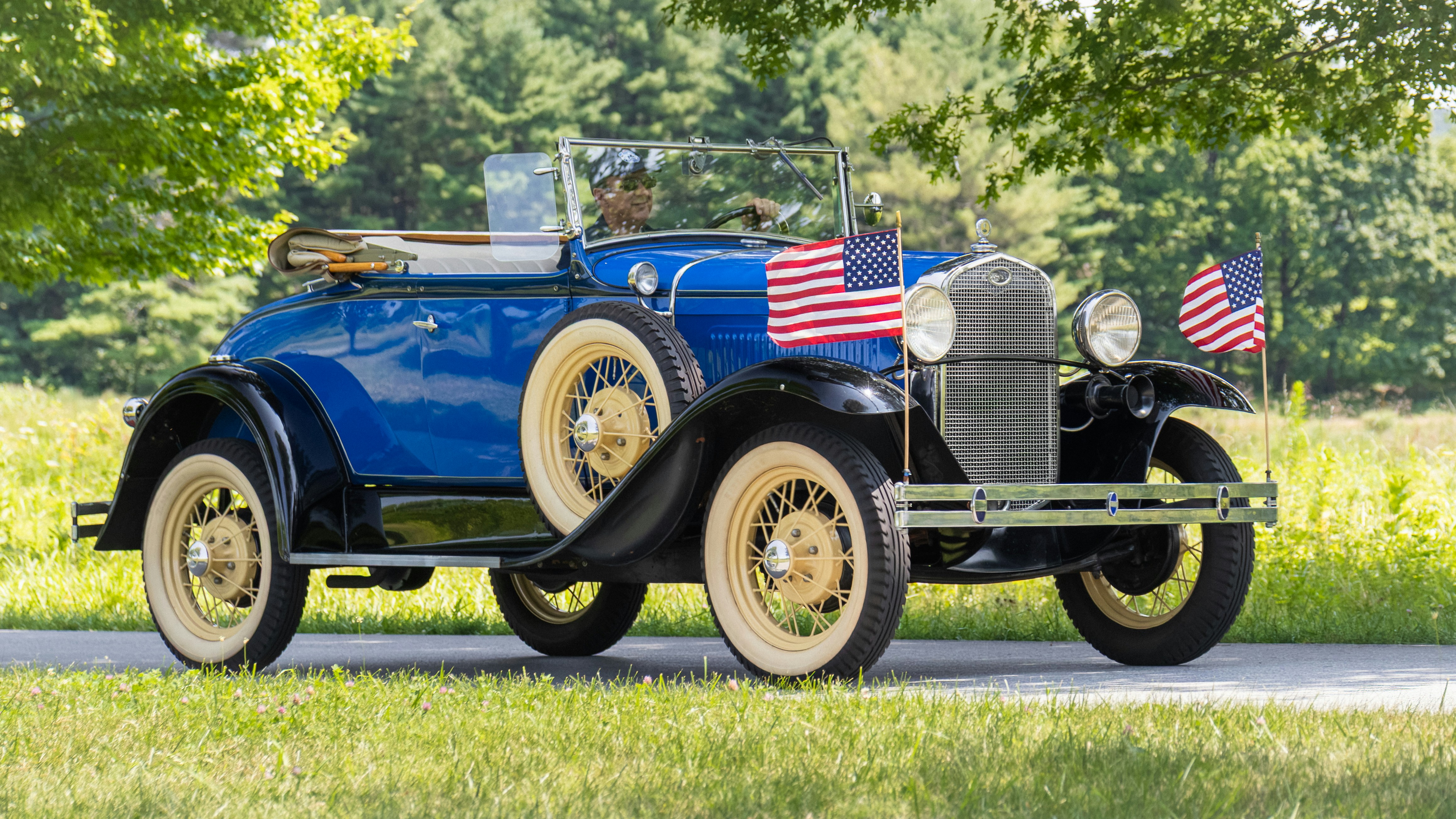 A pristine blue vintage car with beige tires and American flags on a sunlit roadside, emphasis on its chrome details and classic lattice grille.