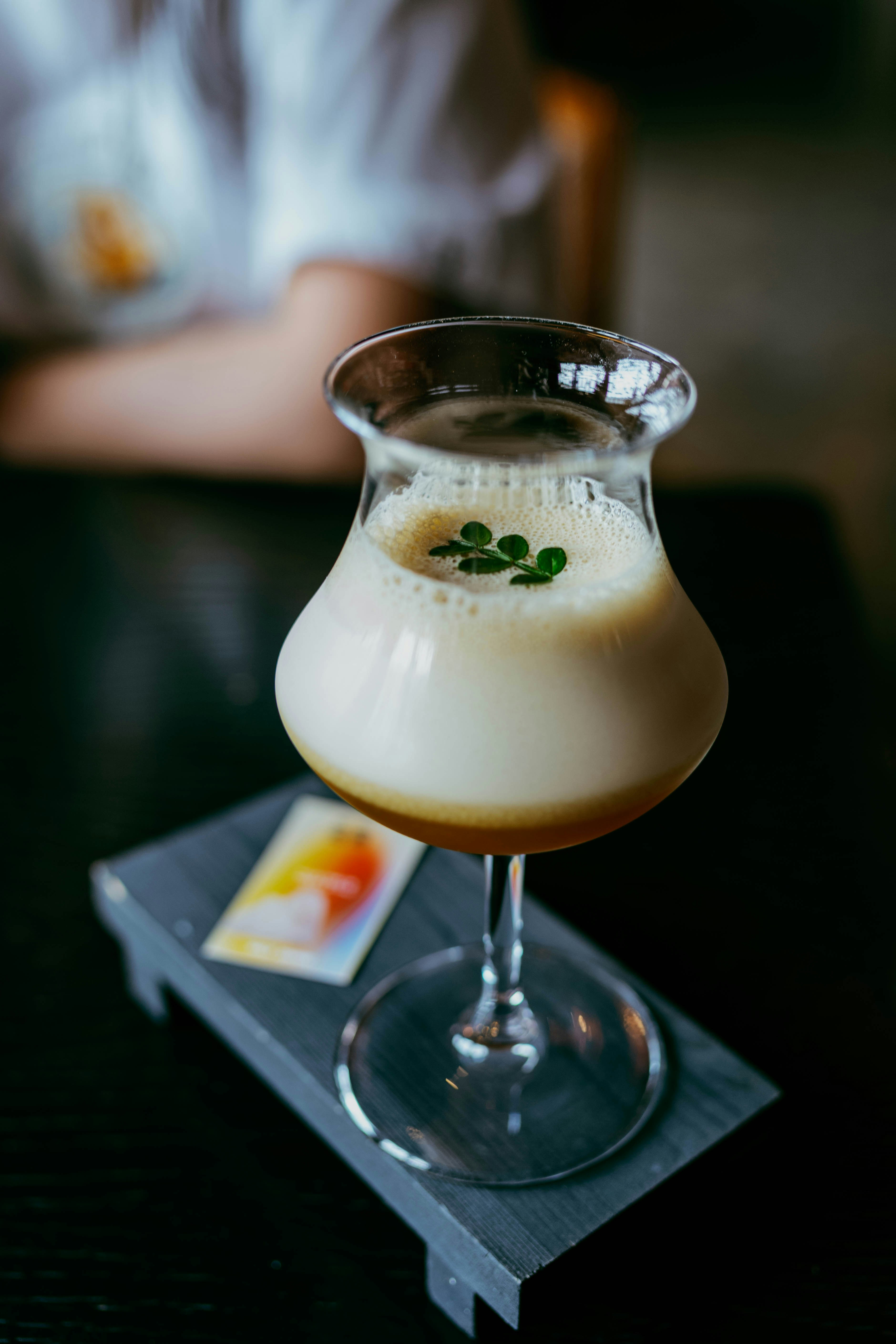 Creamy cocktail in a tulip glass with a foamy head sits on a dark surface, a small garnish visible through the glass. A card and blurred background suggest a bar or cafe setting.