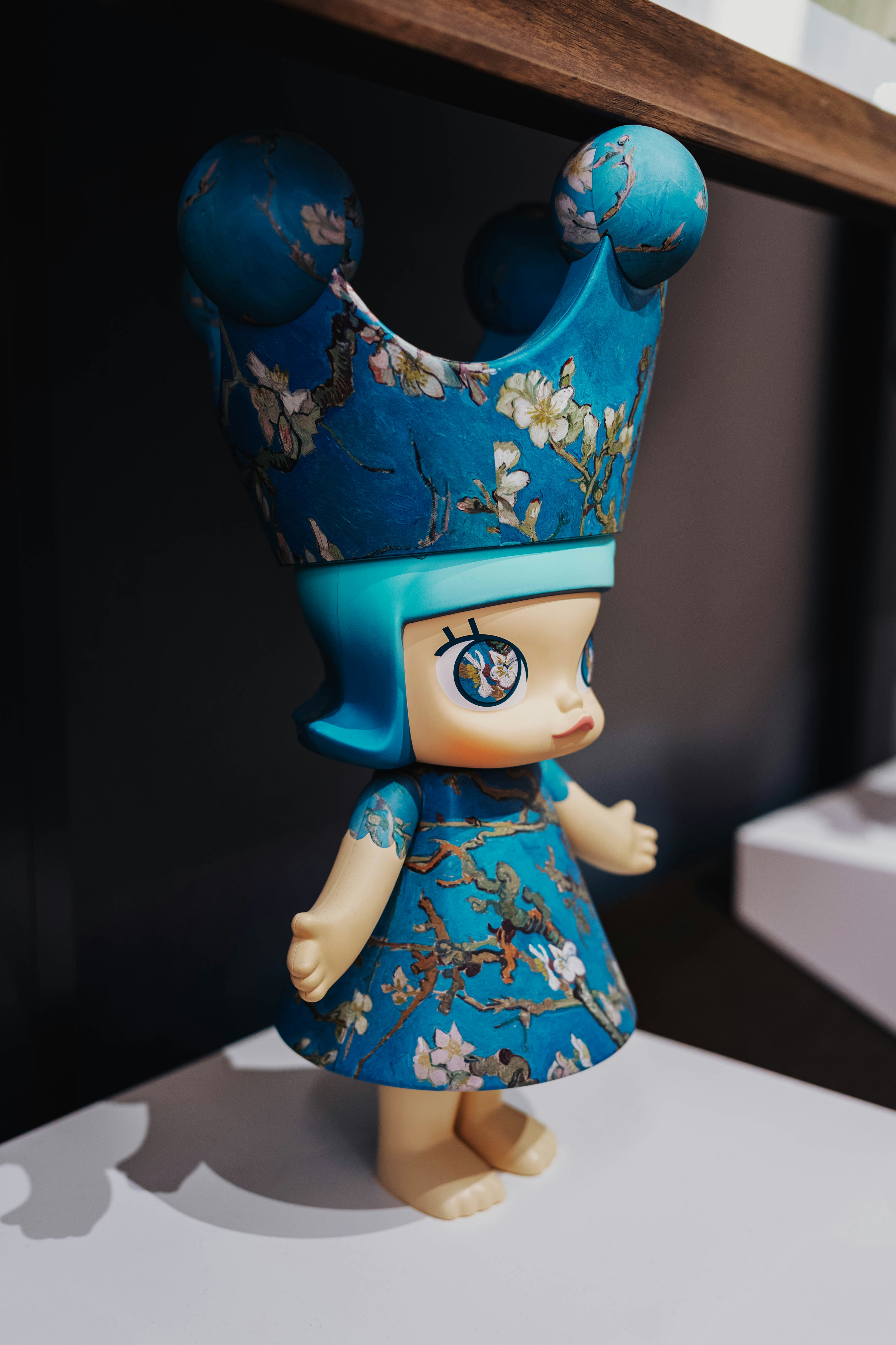 A blue floral toy figure stands on a white surface with a dark background, its oversized crown-like hat drawing the eye.