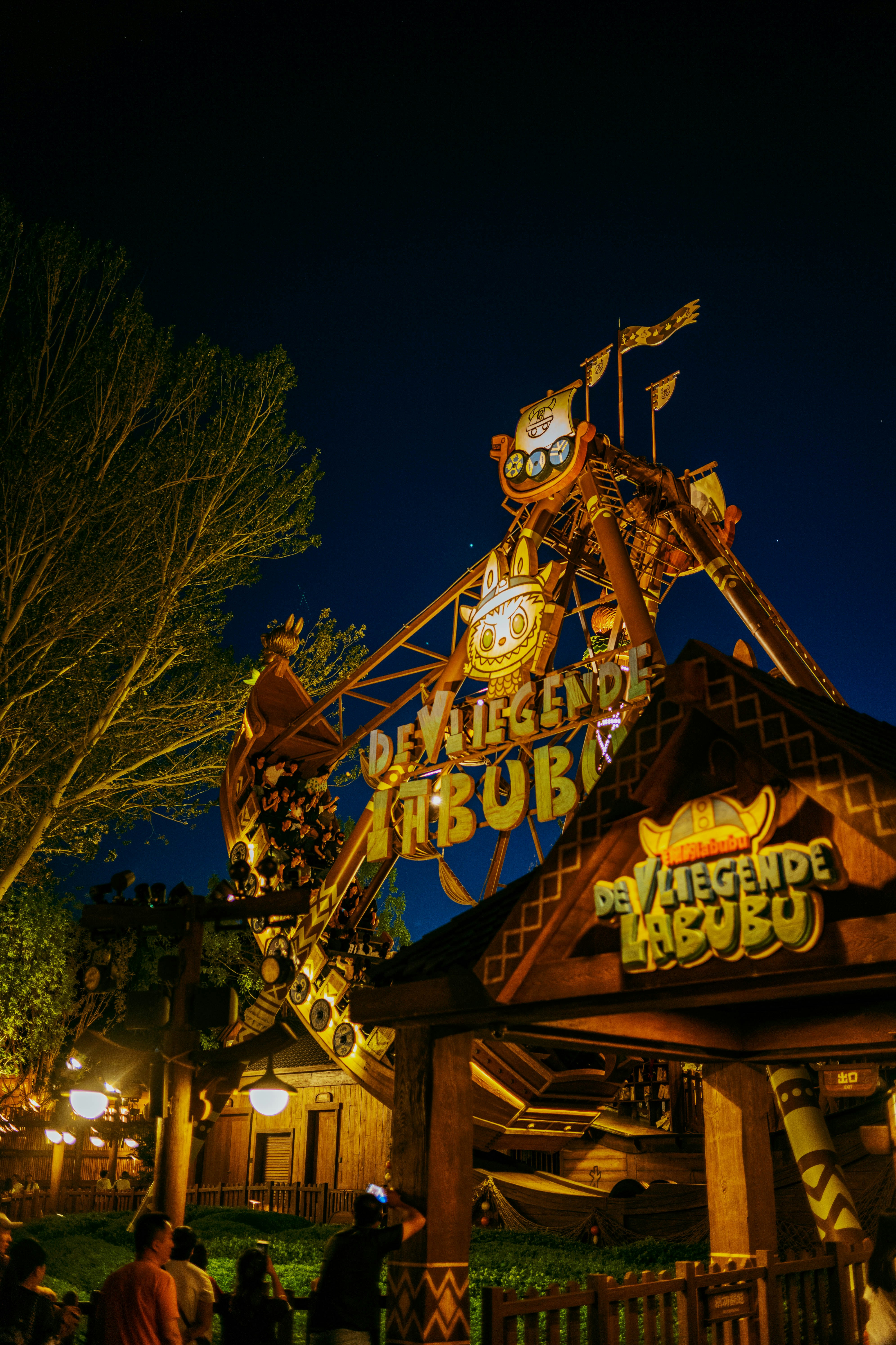 Night scene of a lit carnival ride with carved wooden facade and illuminated signage, a lively entrance crowd in the foreground. This is a theme-park attraction image.
