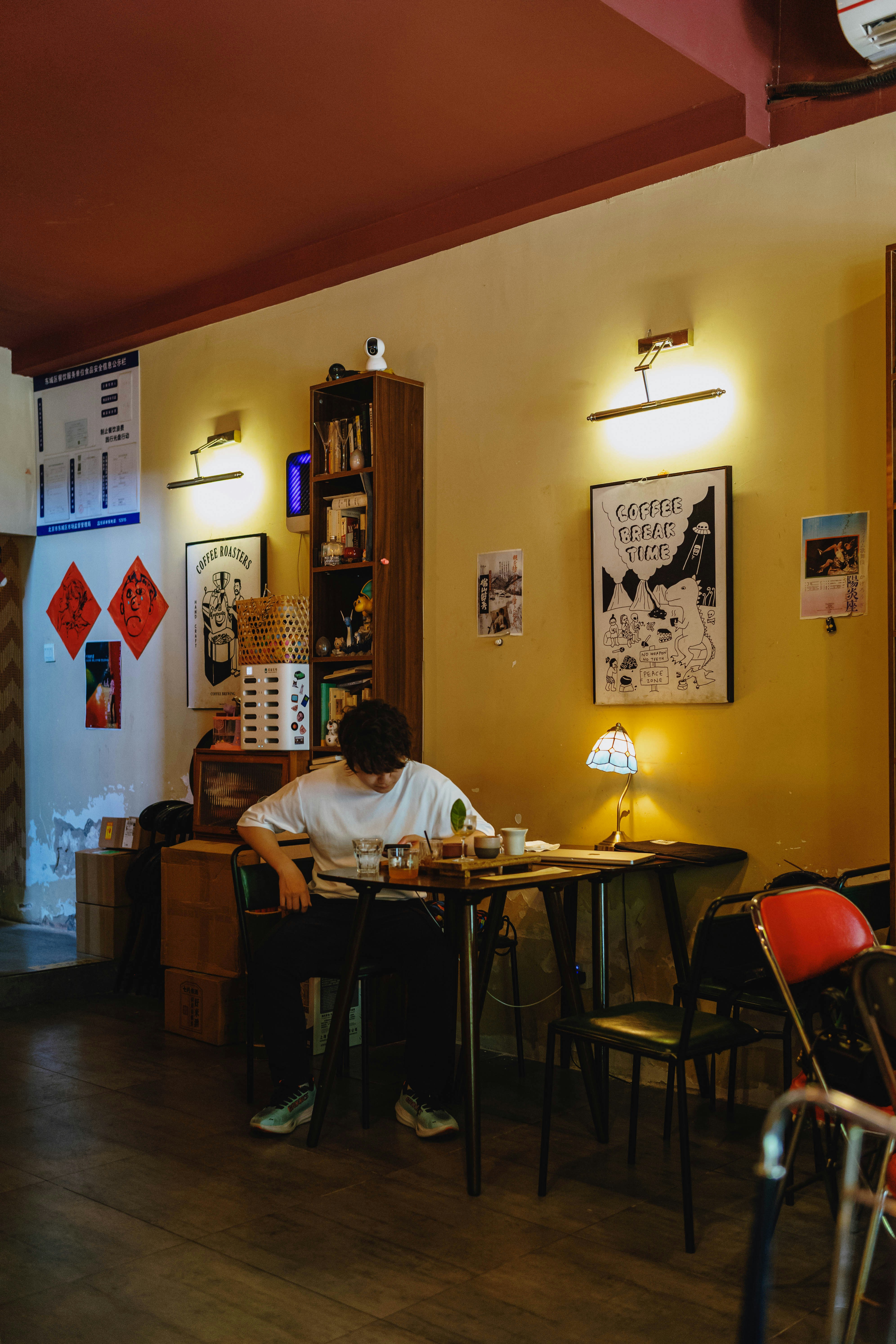 A solitary patron sits at a table in a warmly lit café, surrounded by shelves, posters, and a small lamp, conveying a calm, intimate moment within a cozy interior. The scene includes vintage furniture and subtle color tones.