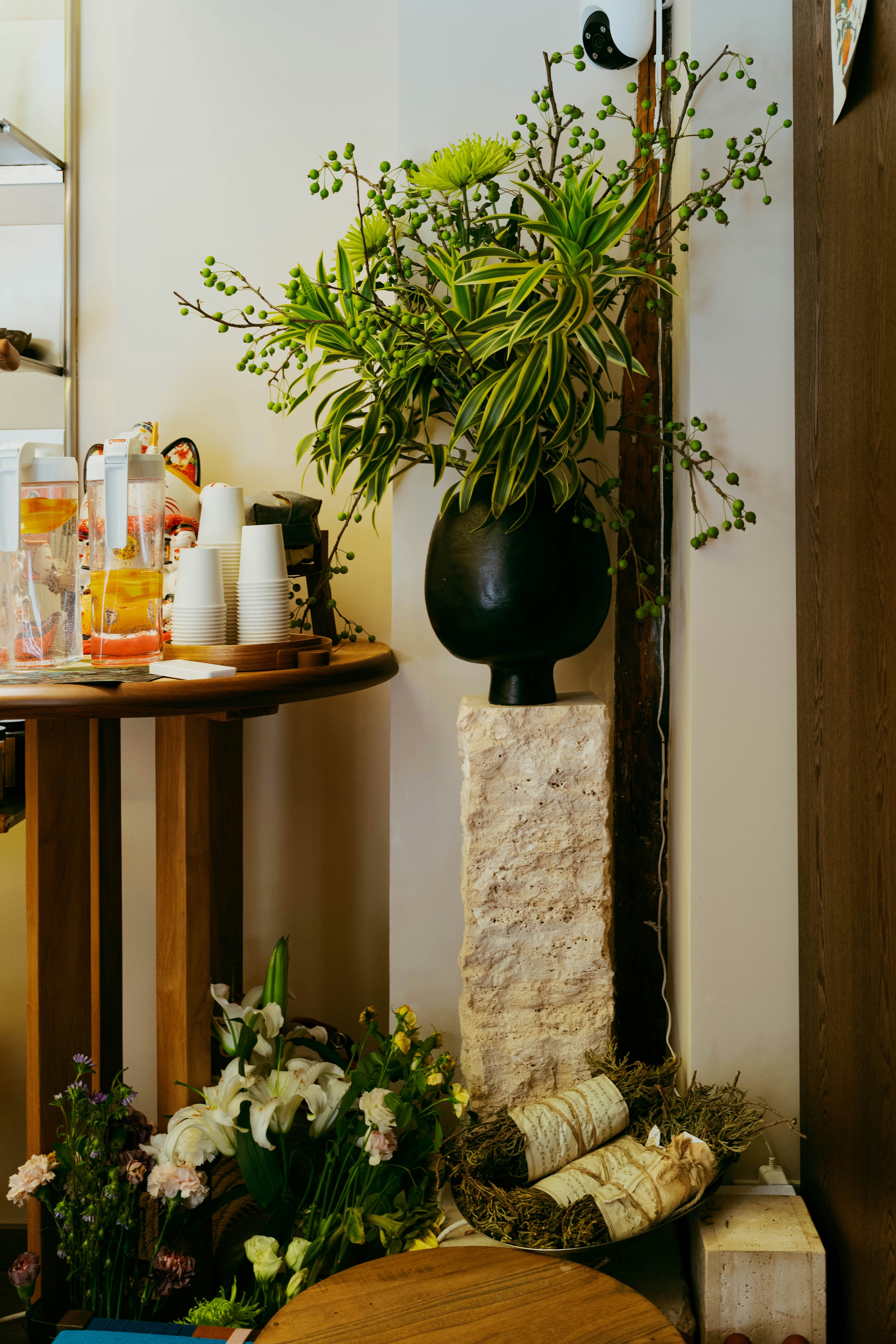 A still life corner with a lush potted plant atop a stone pedestal, surrounded by easterly light, a wooden table with cups and beverages, and a base of white flowers and foliage.