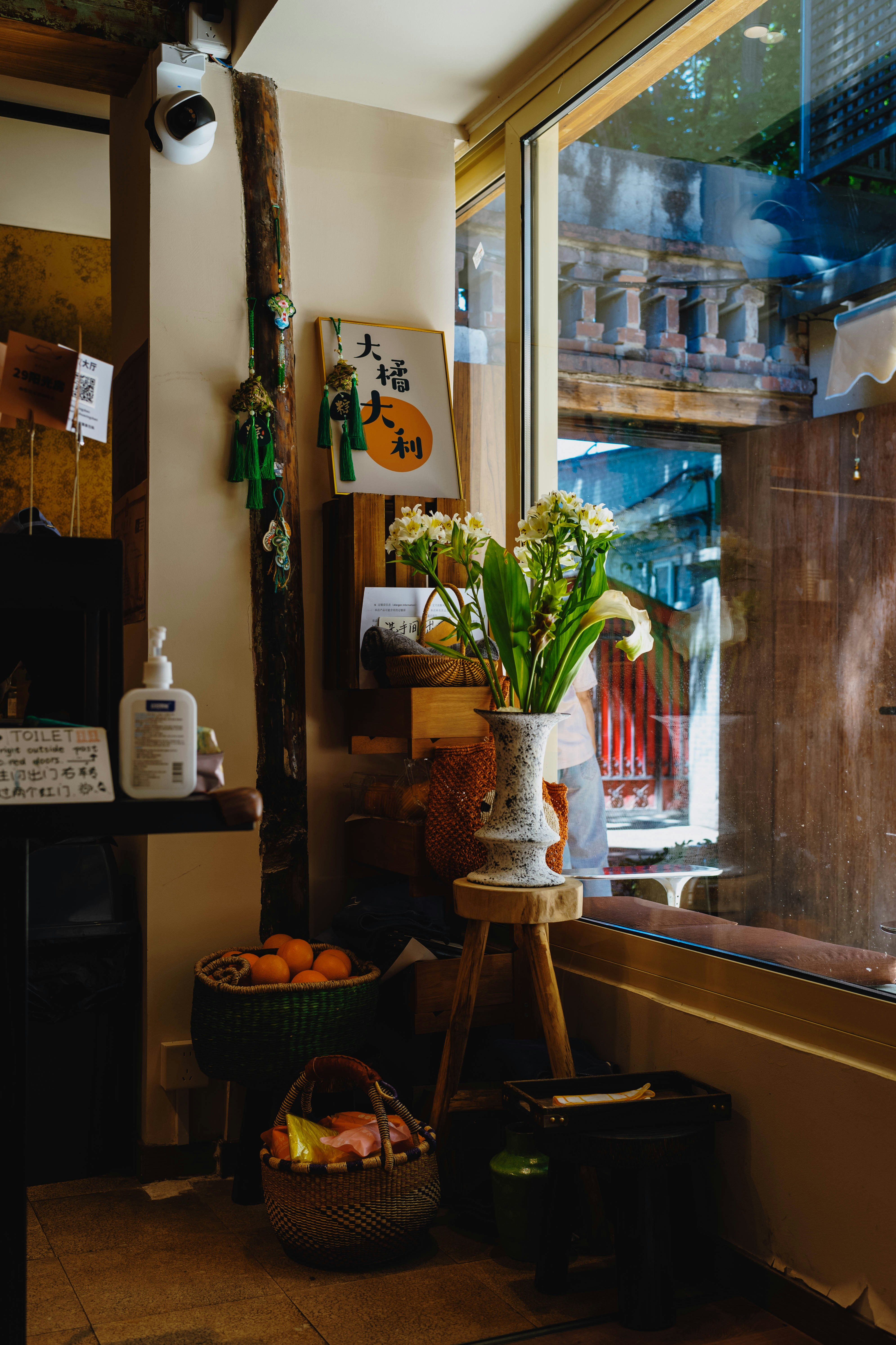 A still life inside a cozy shop features a white vase with fresh flowers on a wooden stand beside a bright window, with baskets of fruit and everyday items nearby. A soft, warm glow fills the scene, highlighting texture and color nuances.