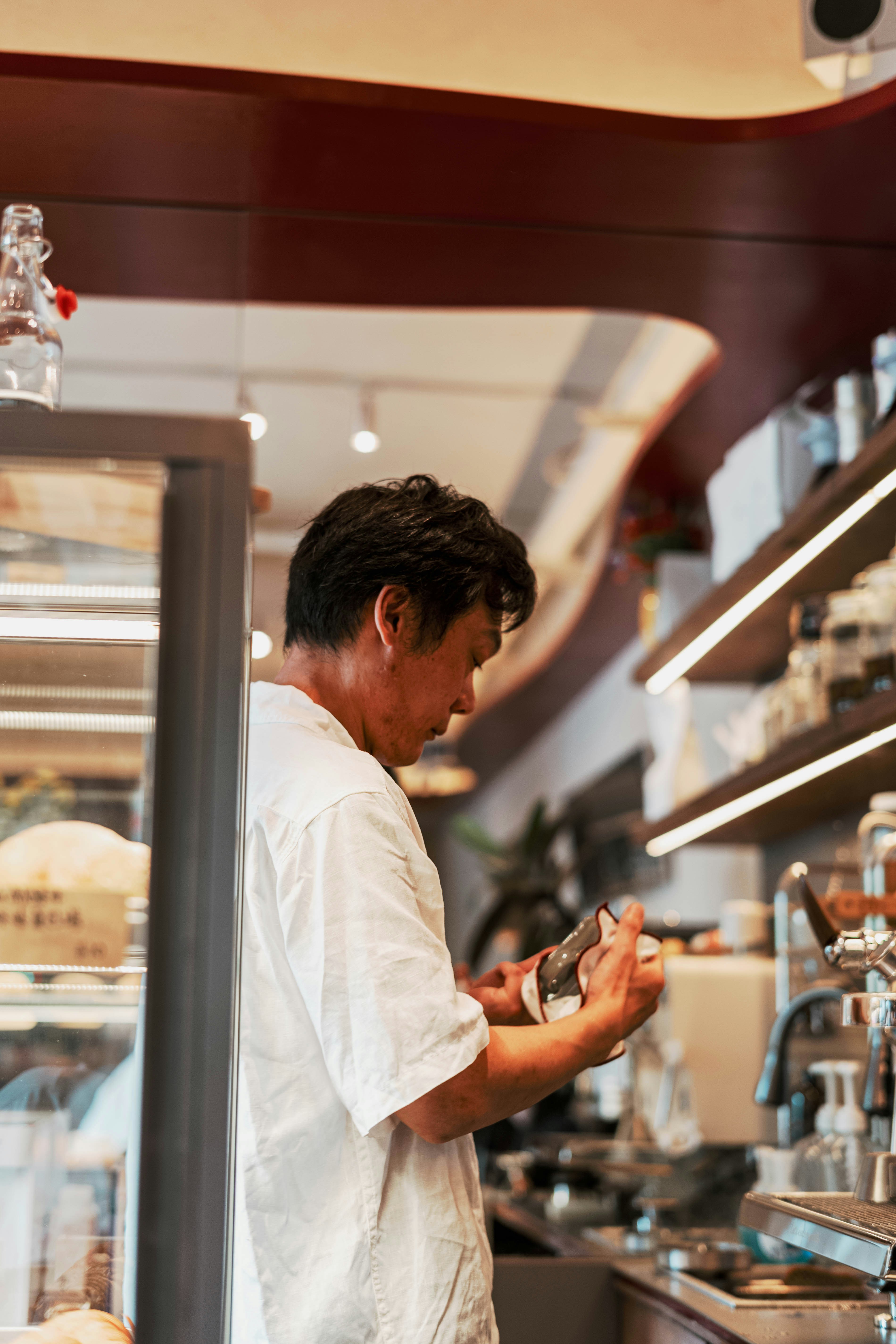A focused portrait of a man in a white shirt working behind a café counter, with beverage equipment and warm lighting in the background. The scene emphasizes the craft environment and the barista’s activity.