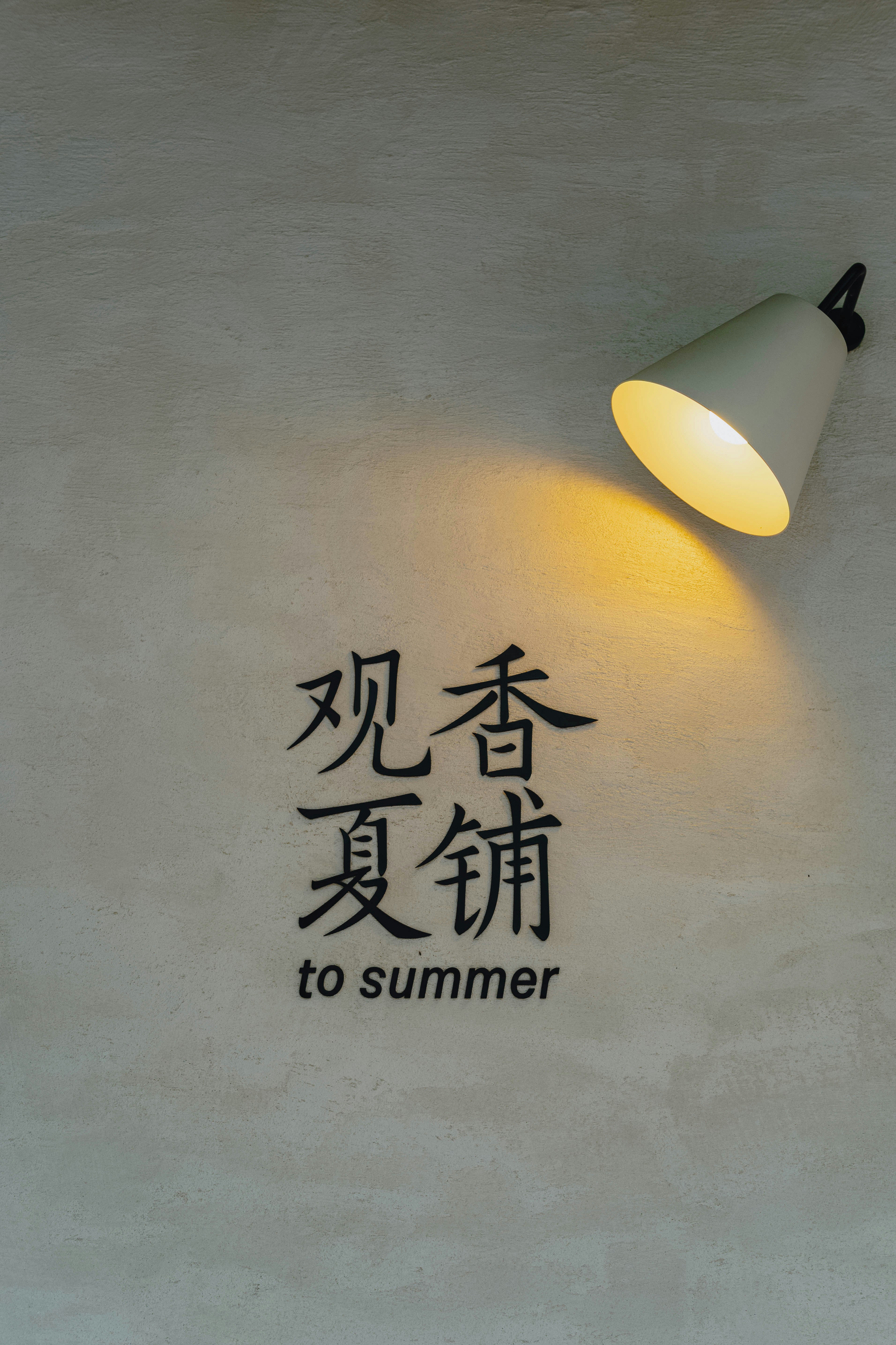 A wall-mounted lamp casts a warm pool of light onto a vertical signboard featuring Chinese characters and the English phrase to summer, creating a soft, inviting ambiance.