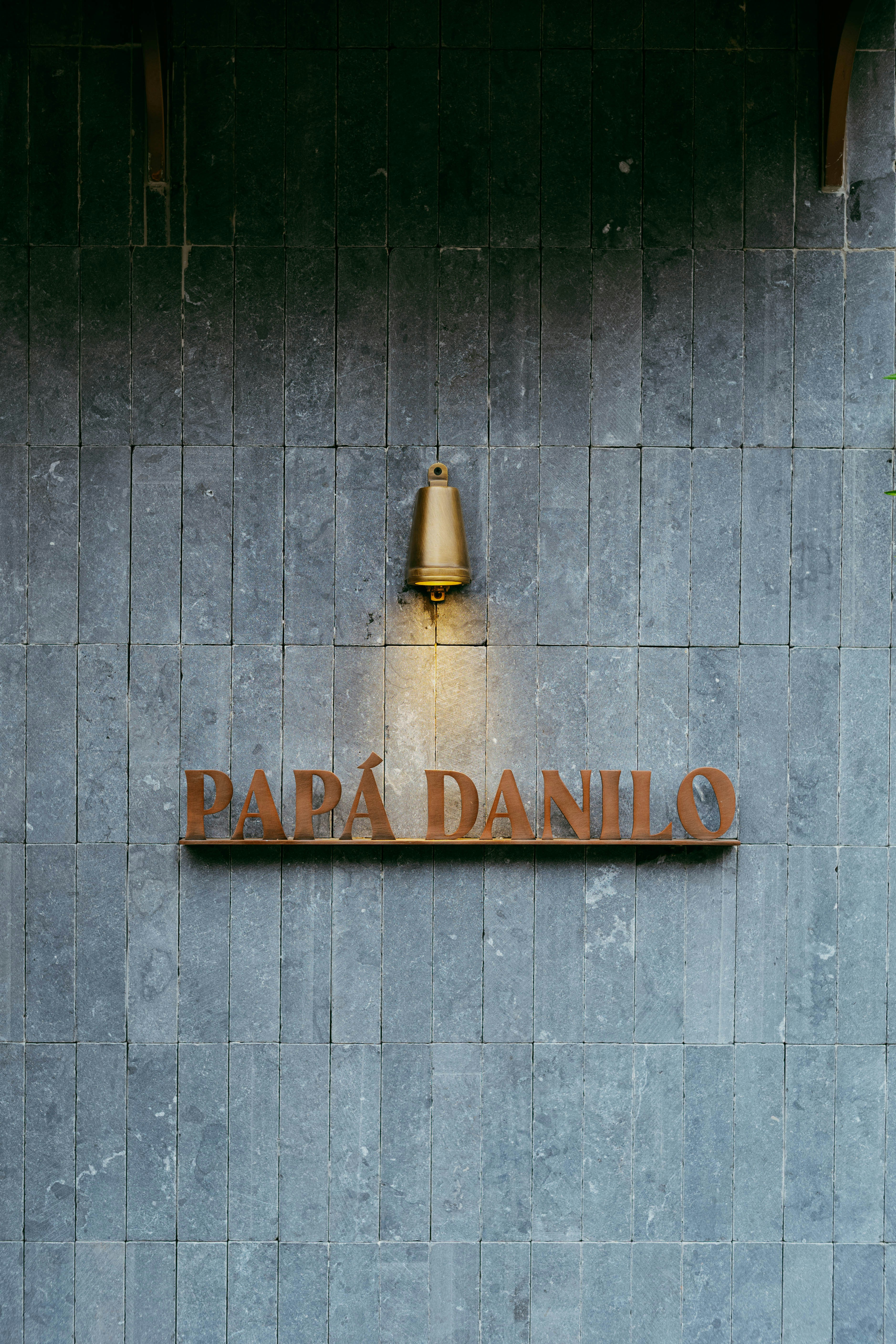 Weathered blue tile wall with a single warm wall lamp above a metal sign that reads PAPÁ DANILO, centered and softly lit to draw the eye downward along the texture of the surface.