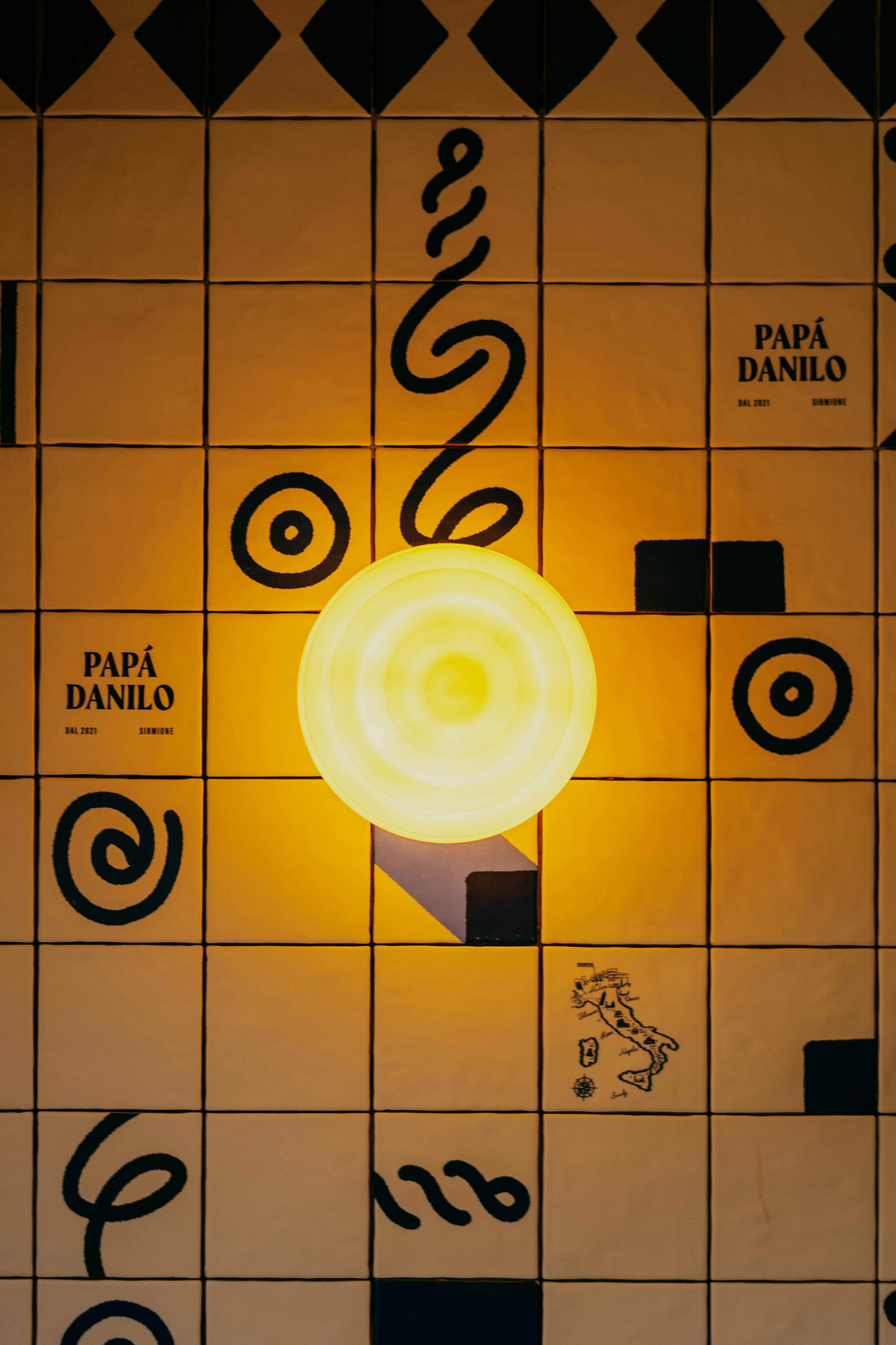 Warm-lit circular lamp suspended against a tiled wall filled with abstract symbols and logos; a modern, decorative scene highlighting graphic geometry and ambient glow.