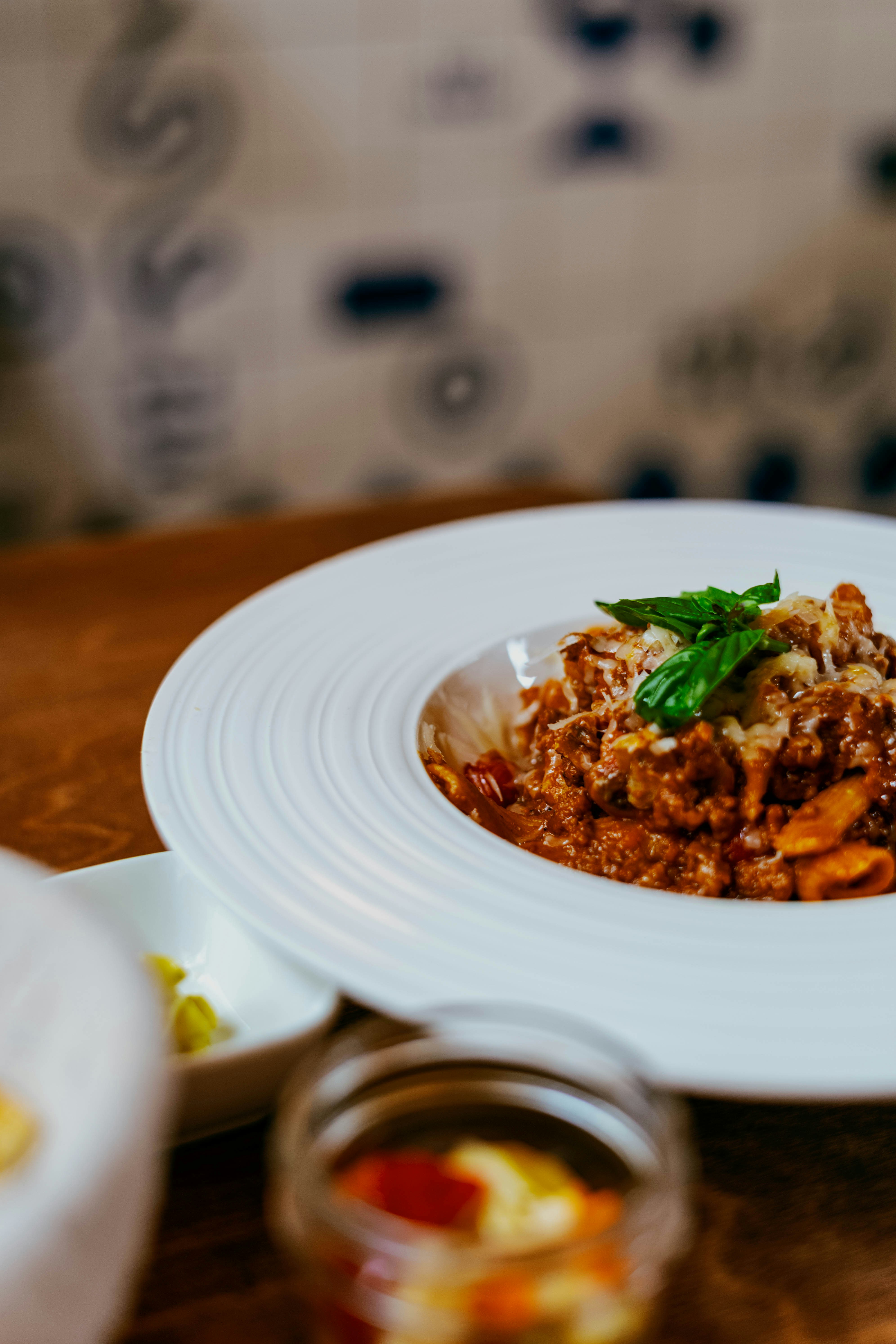 A plated pasta dish with a rich sauce and fresh basil sits on a white, concentric-ring plate; a small jar and side bowls are blurred in the foreground, suggesting a dining setting.