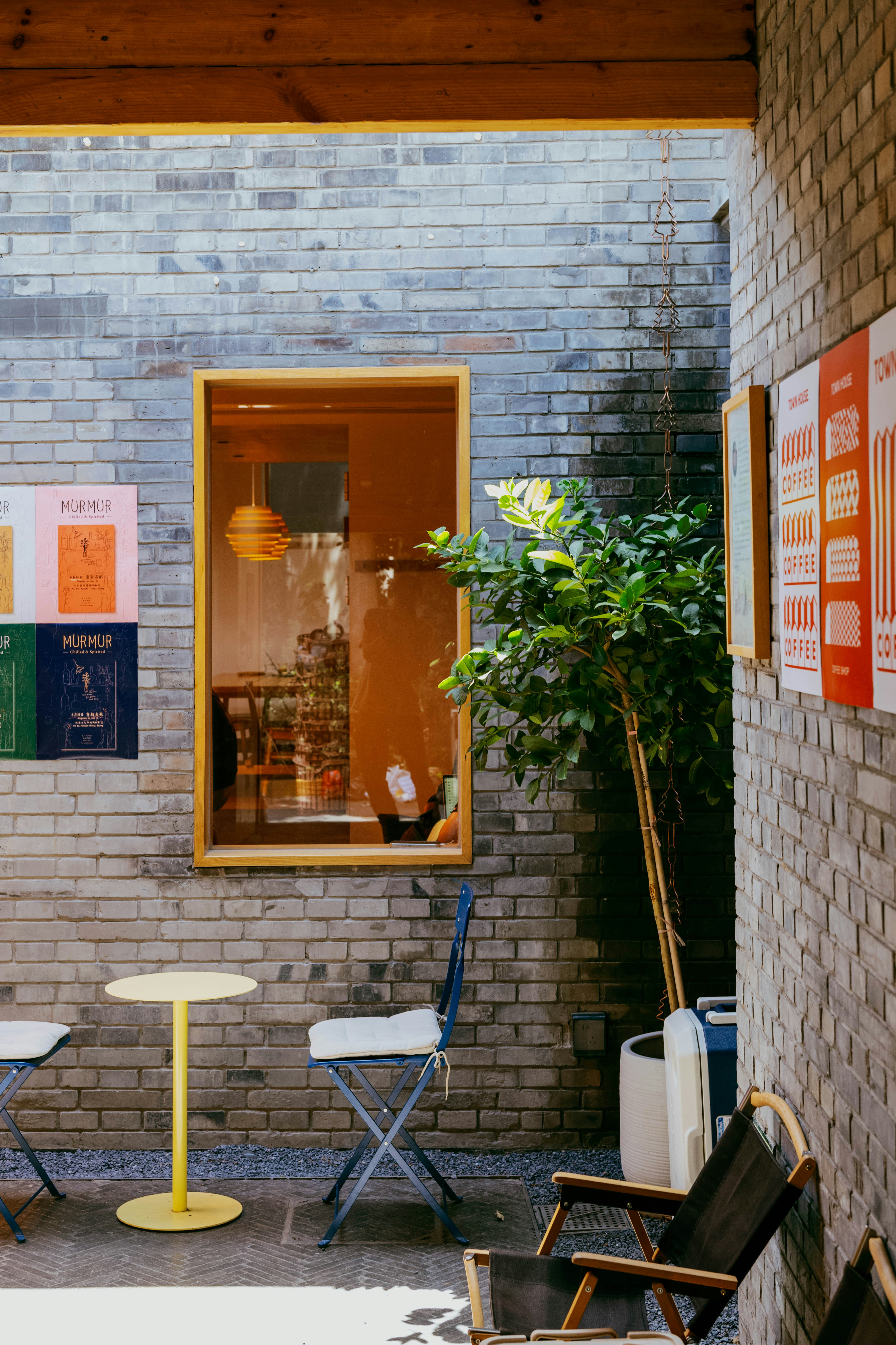 A small outdoor seating area beside a brick wall with a yellow-framed window, colorful posters, and a potted plant casting soft shade over chairs and a yellow table.