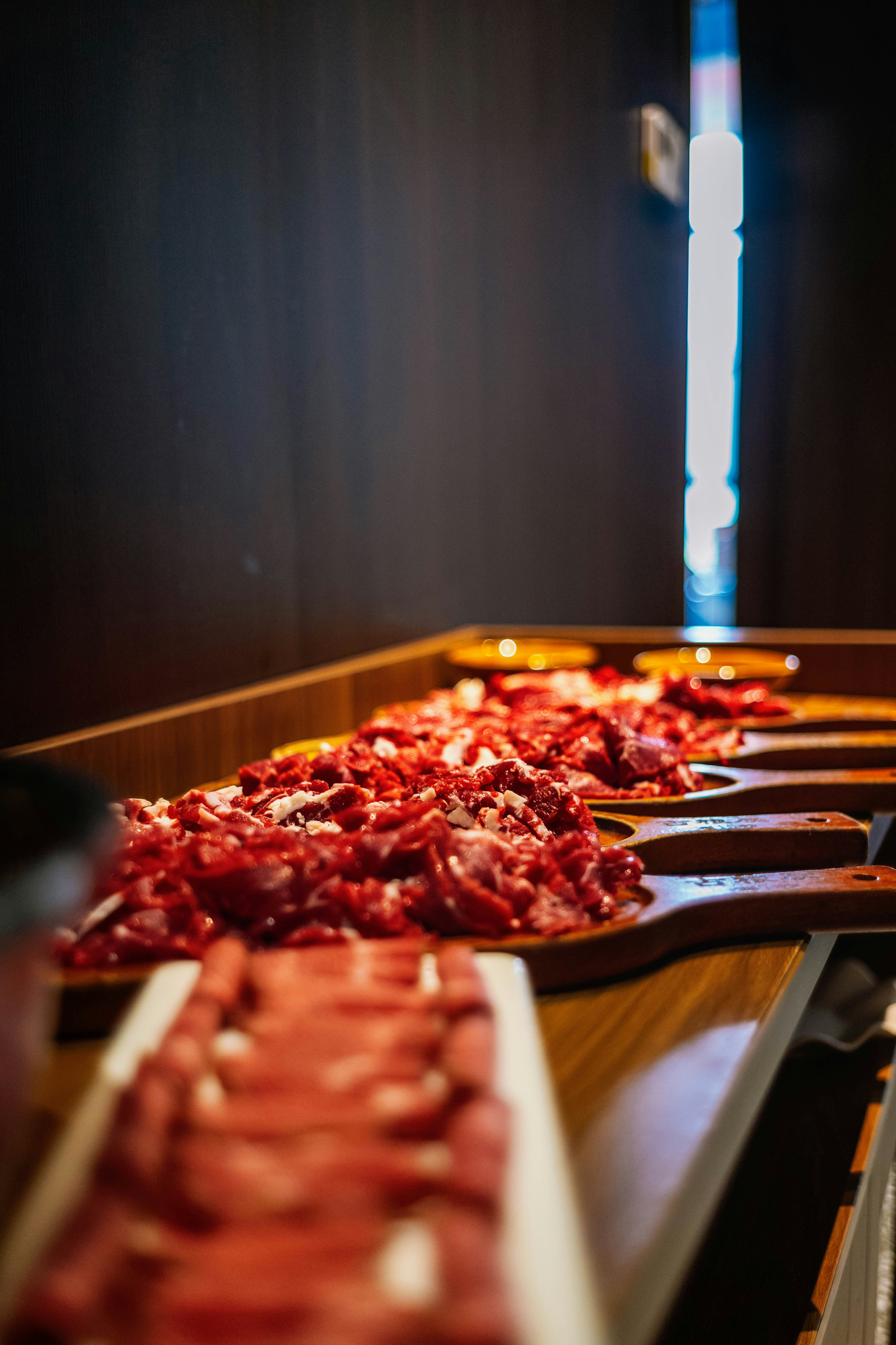 Sliced meats arranged on wooden boards under dim, neon-lit background; a close-up of raw cuts emphasizing color and texture. A food-prep scene with a focus on the foreground meat trays.
