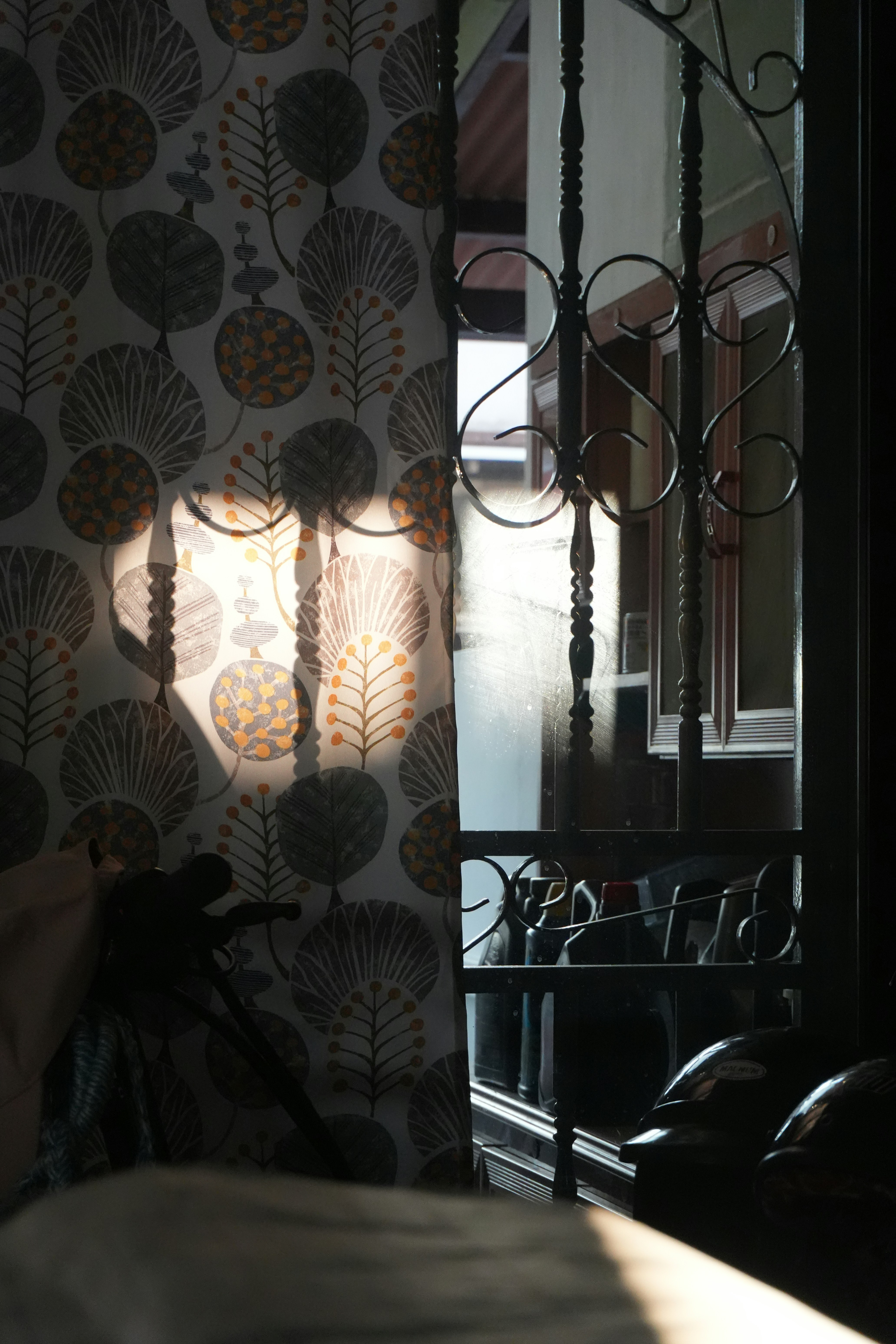Dim interior with patterned wallpaper on left and a barred window/door on the right; sunlight creates a soft highlight on the wall and foreground objects in shadow.