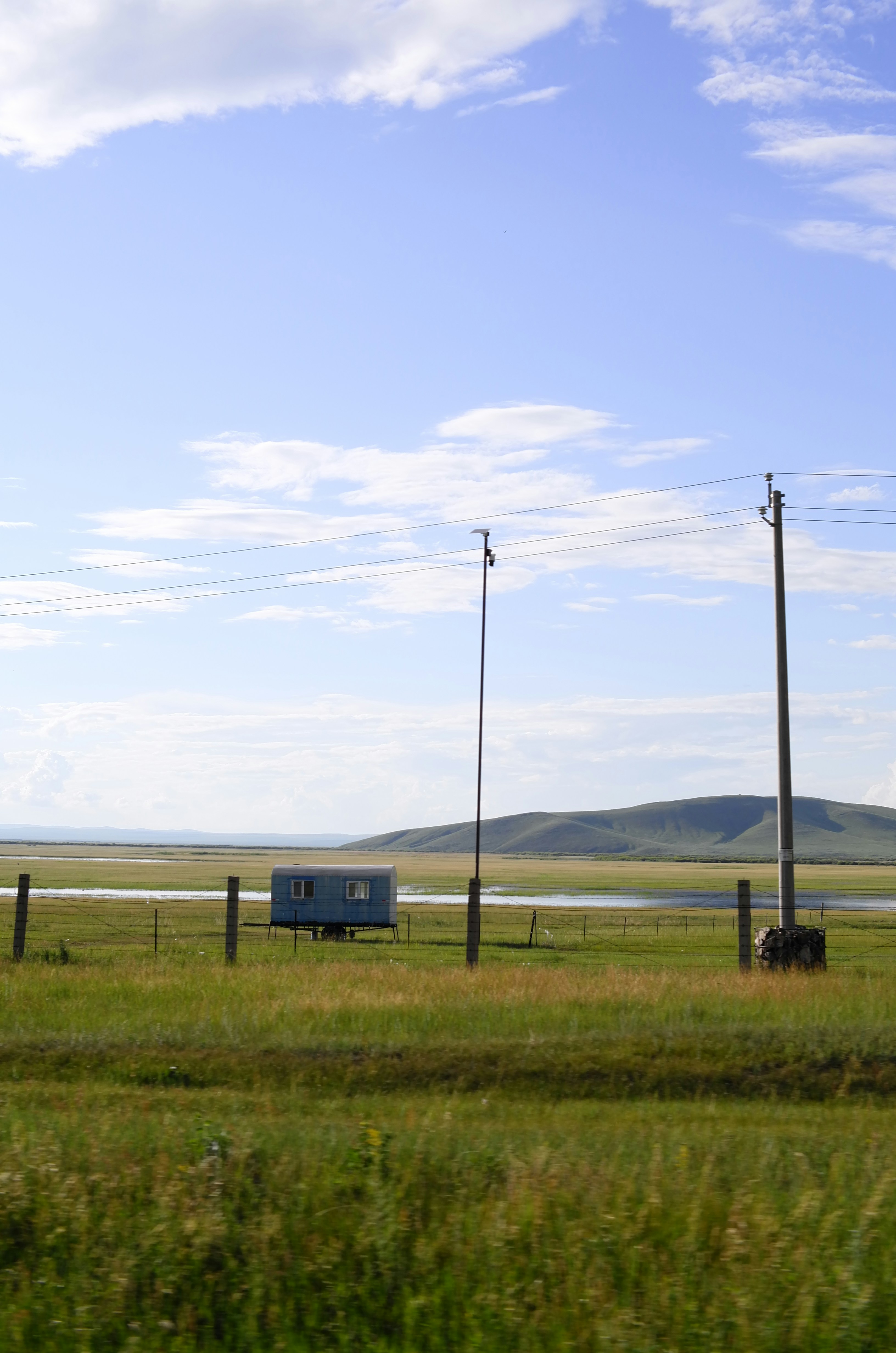 Small blue cabin sits beside a fence under a bright sky, with distant hills and utility poles creating a rural landscape.