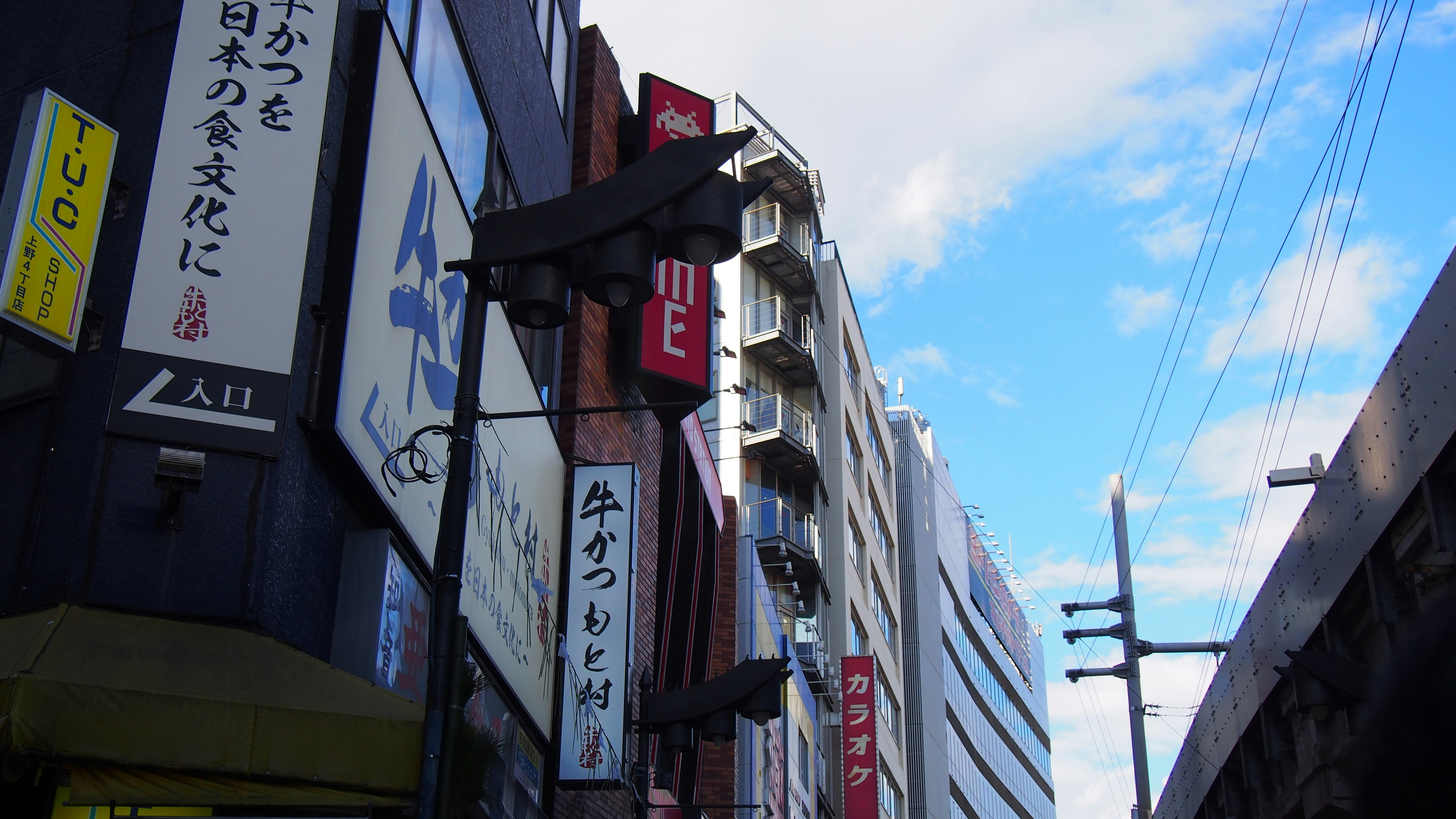 Bustling alley with tall buildings lined by vertical neon signs and a bright blue sky above.
