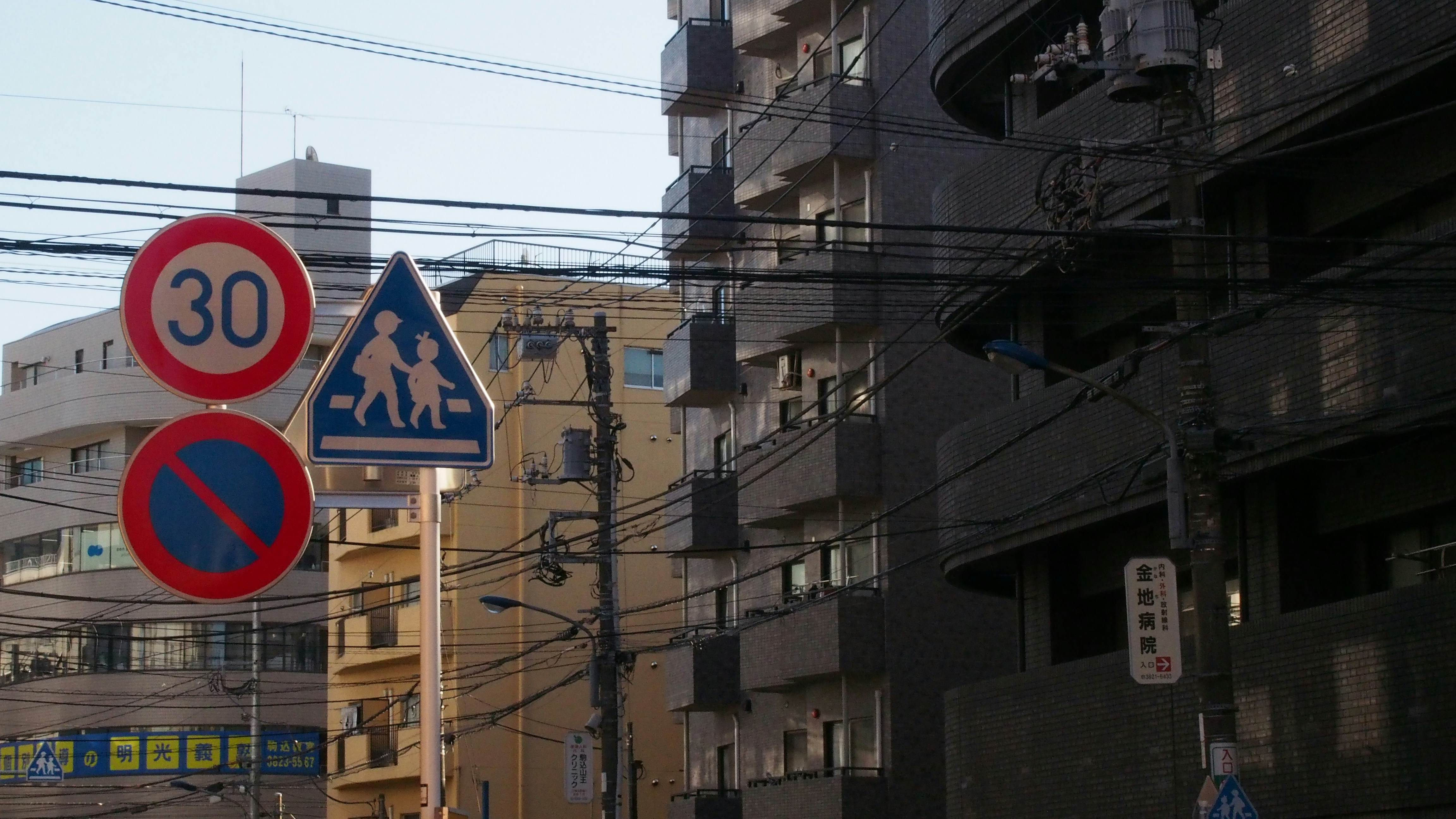 A dense city street scene featuring prominent traffic and pedestrian signs with apartment buildings in the background. The composition emphasizes urban signage and overhead utility lines.