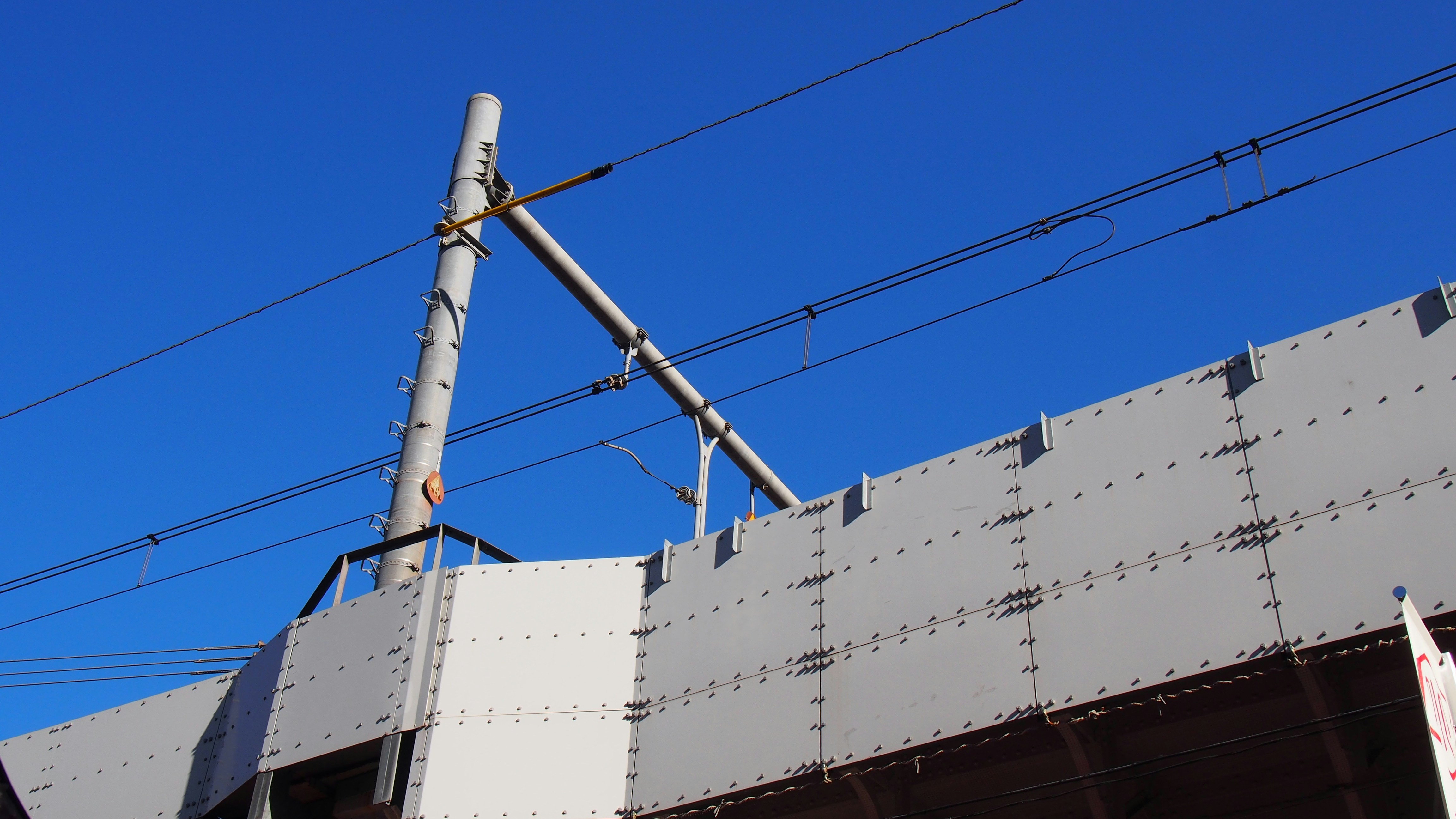 Industrial scaffolding and overhead wiring rise against a vivid blue sky, capturing a minimalist urban structure and the texture of riveted metal panels.