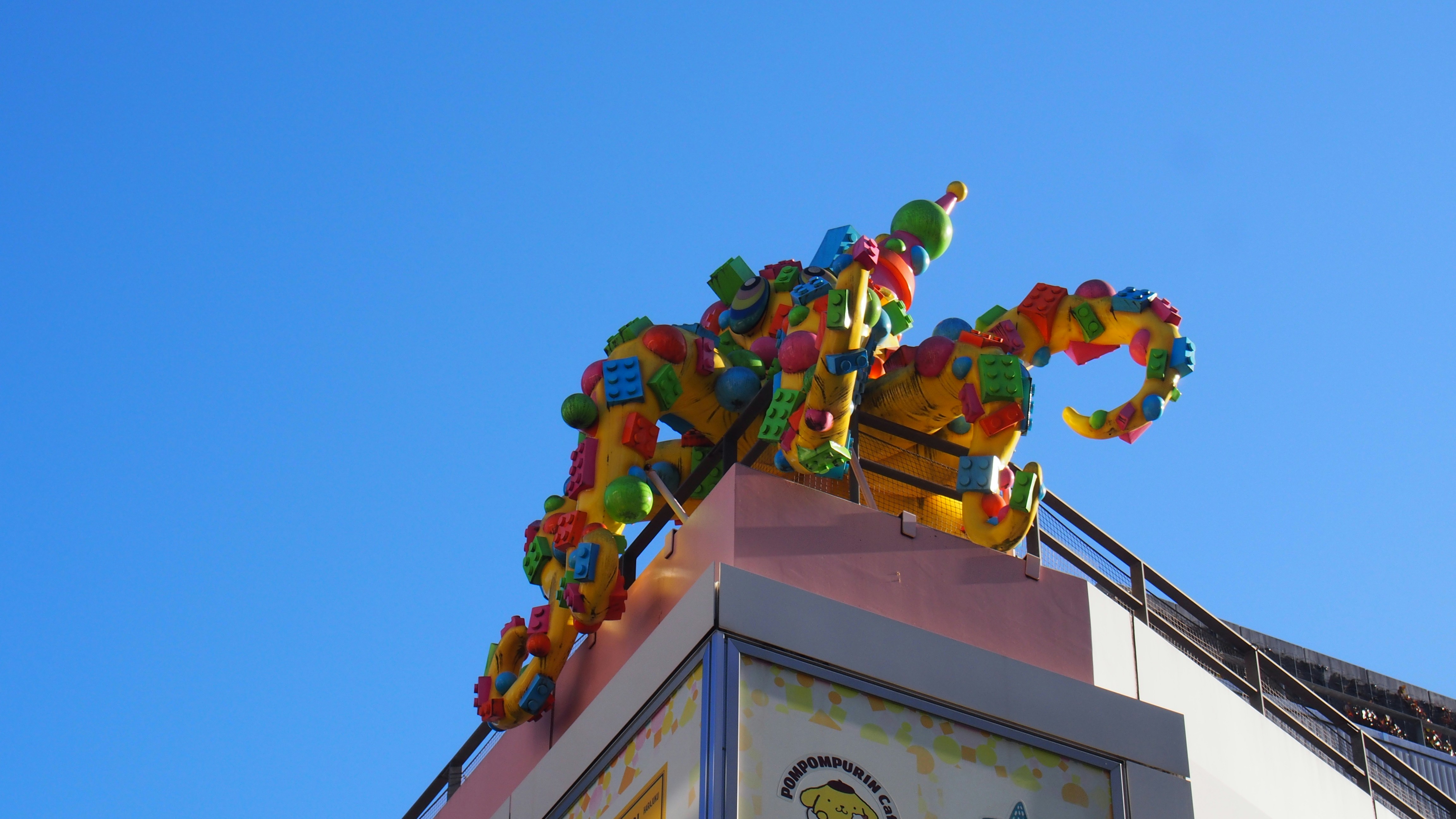 Vivid ornamental figures atop a sloped roof against a bright blue sky, with a storefront panel visible below. A decorative piece, not a person, stands as the focal point of the architectural scene.
