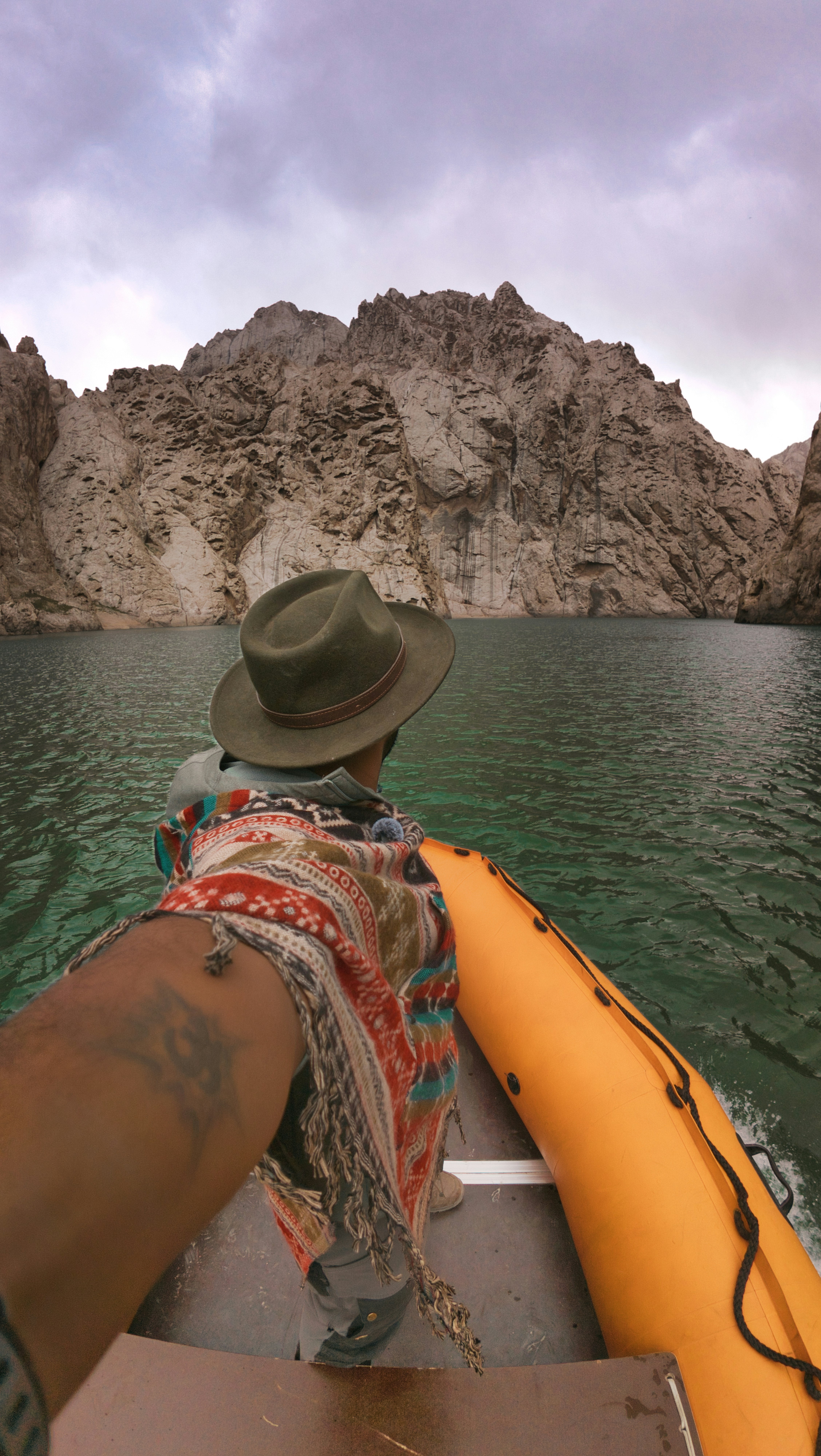 A person in a wide-brim hat sits at the bow of a small boat, facing a rugged canyon wall and green water; colorful scarf and orange inflatable boat create a vivid foreground against a cloudy sky.