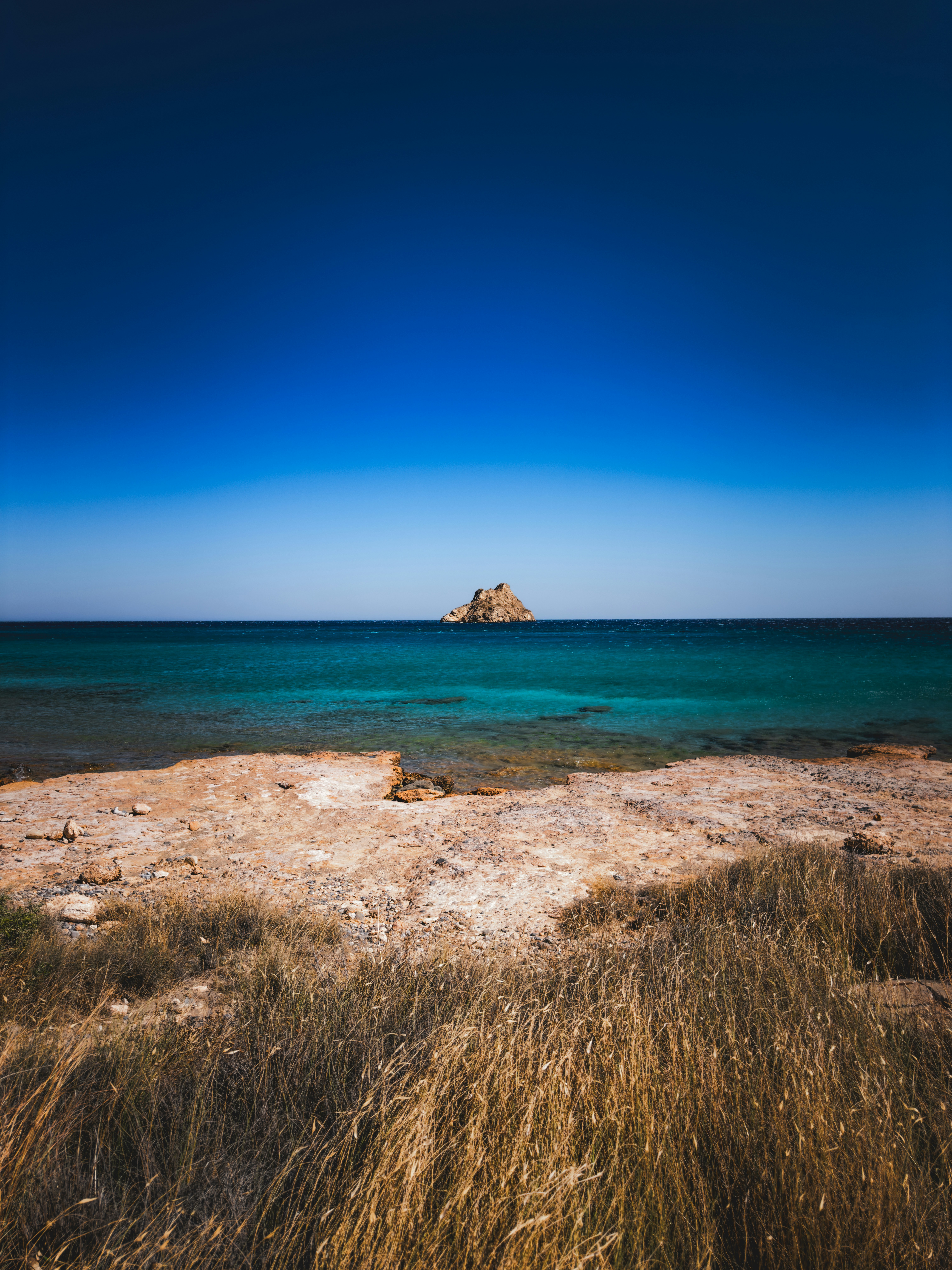 Coastal scene with a small rocky islet centered on the horizon, turquoise sea, and rugged foreground grasses. A serene landscape emphasizing horizon line and natural textures.