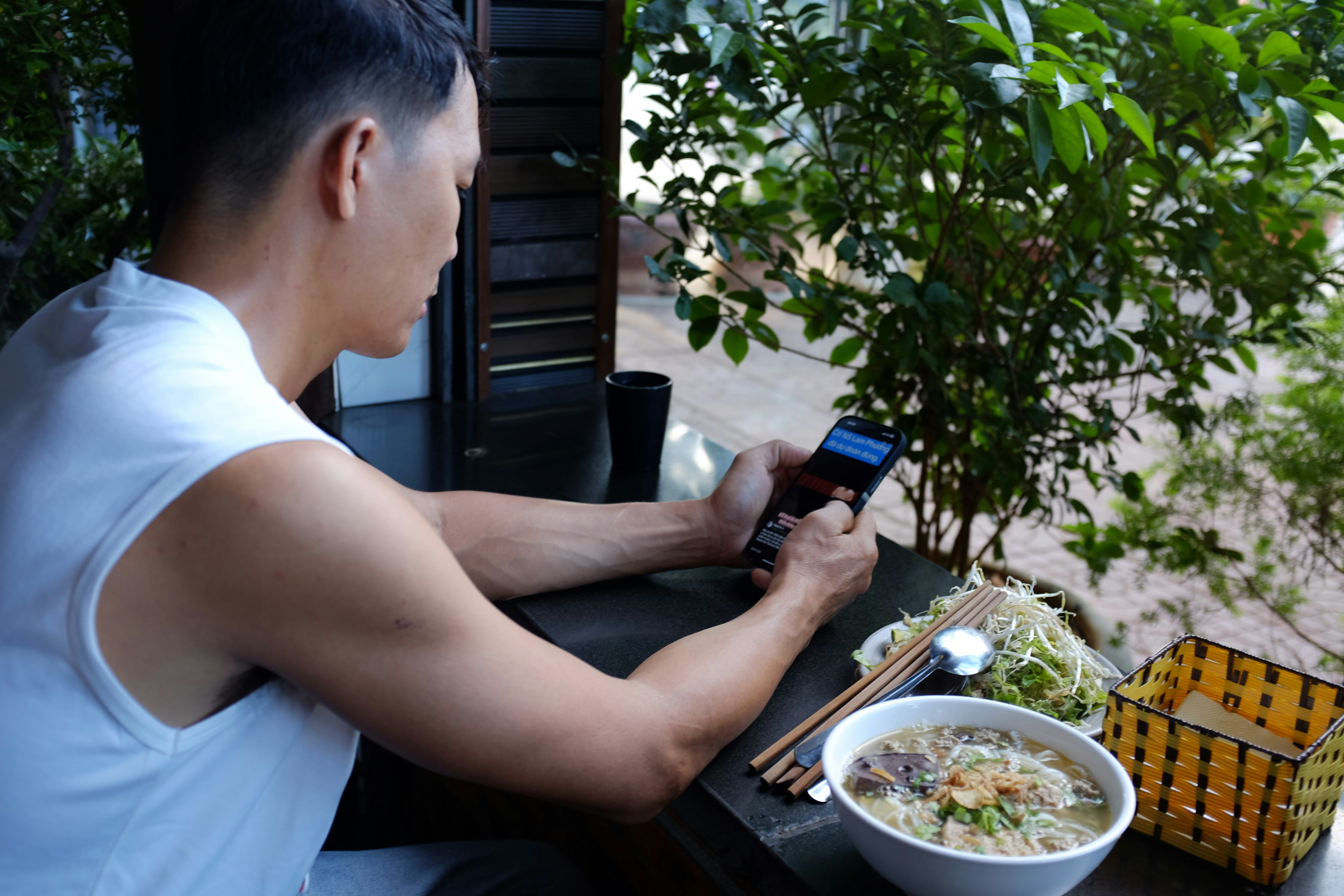 A man in a sleeveless white shirt sits at an outdoor table, focused on a smartphone while a bowl of noodles and chopsticks sit nearby, framed by greenery and a small basket on the table. This is a candid lifestyle scene with a dining setup.
