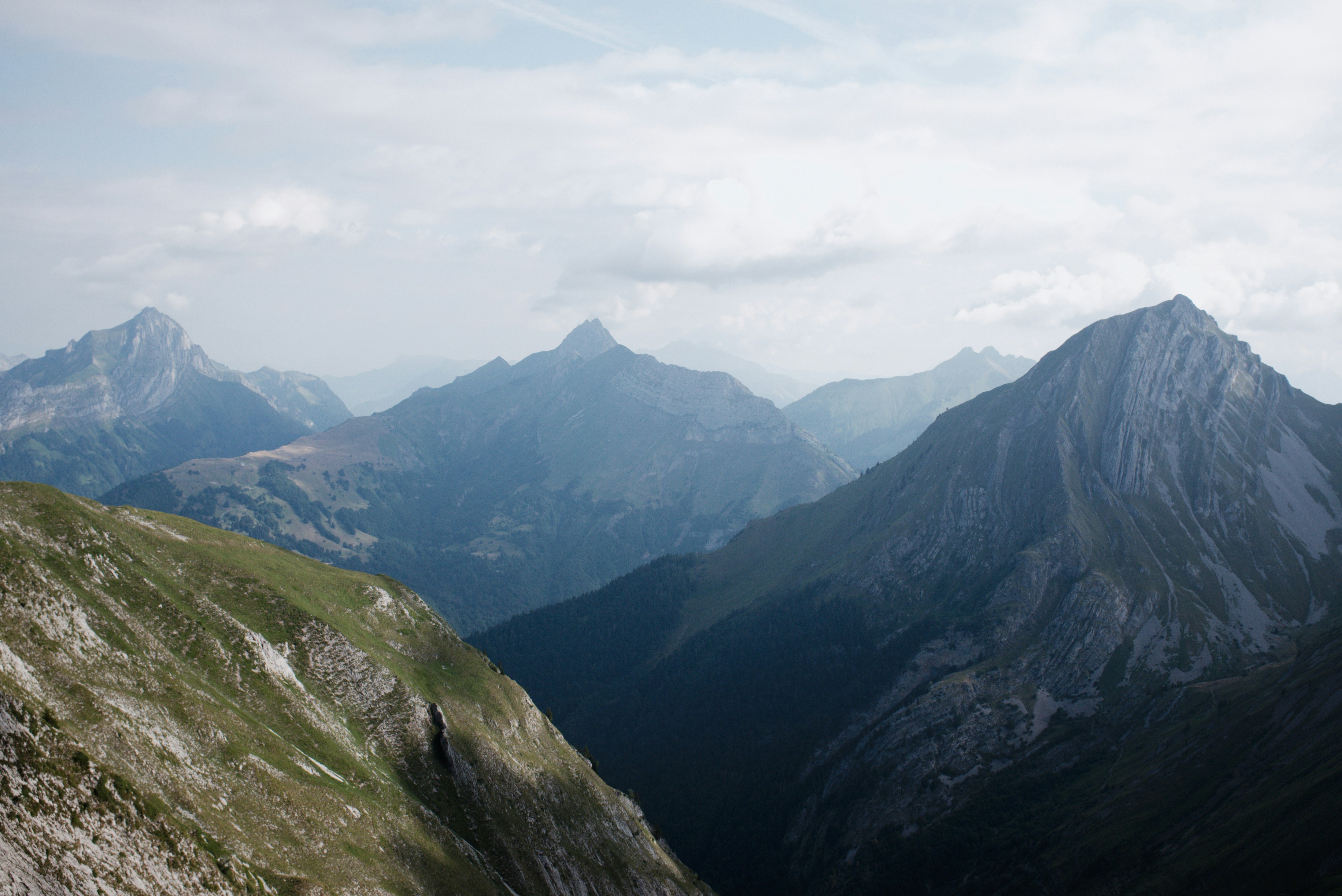 Sinewy mountain ridges stretch into the distance under a pale sky, with a sunlit ledge in the foreground. A sense of grandeur and depth is conveyed by layered ranges and shadowed valleys.