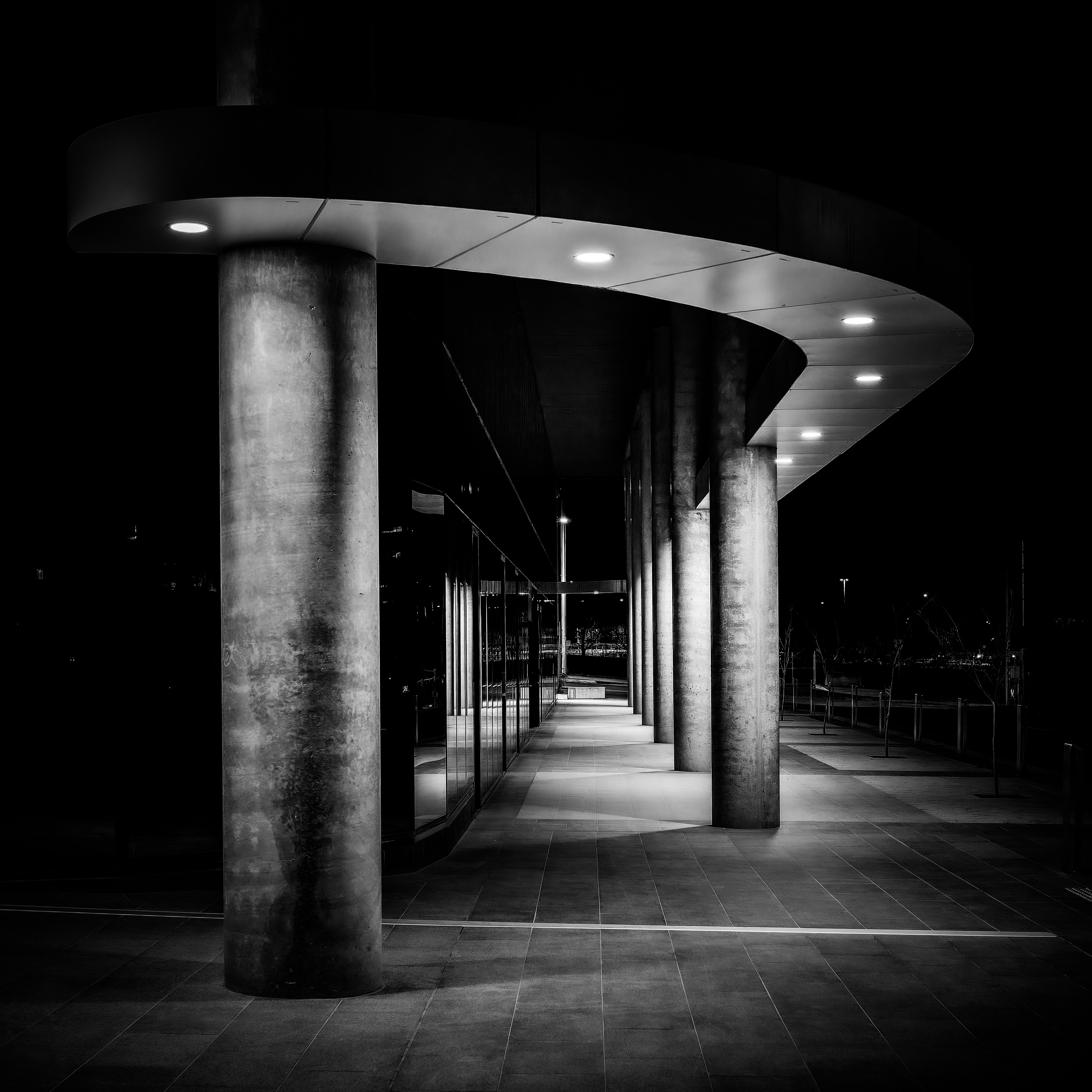 Monochrome night scene of a modern architectural walkway supported by tall concrete columns beneath a curved canopy. The corridor recedes toward a bright vanishing point, emphasizing symmetry and leading lines.