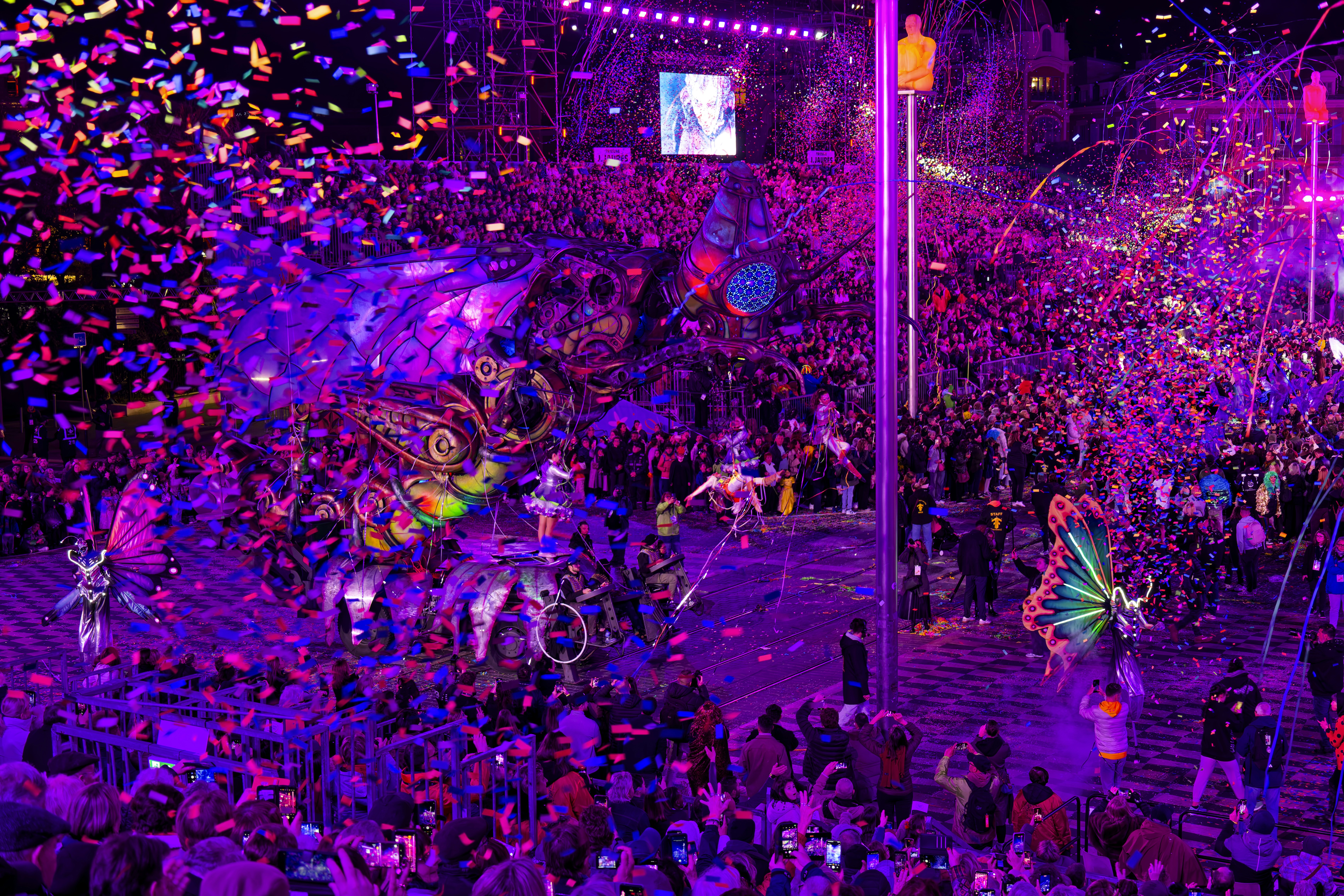 Crowd-filled street carnival bathed in purple and pink neon, with butterfly-wing performers and confetti overhead; vibrant lights and large screens create a festive, immersive scene.