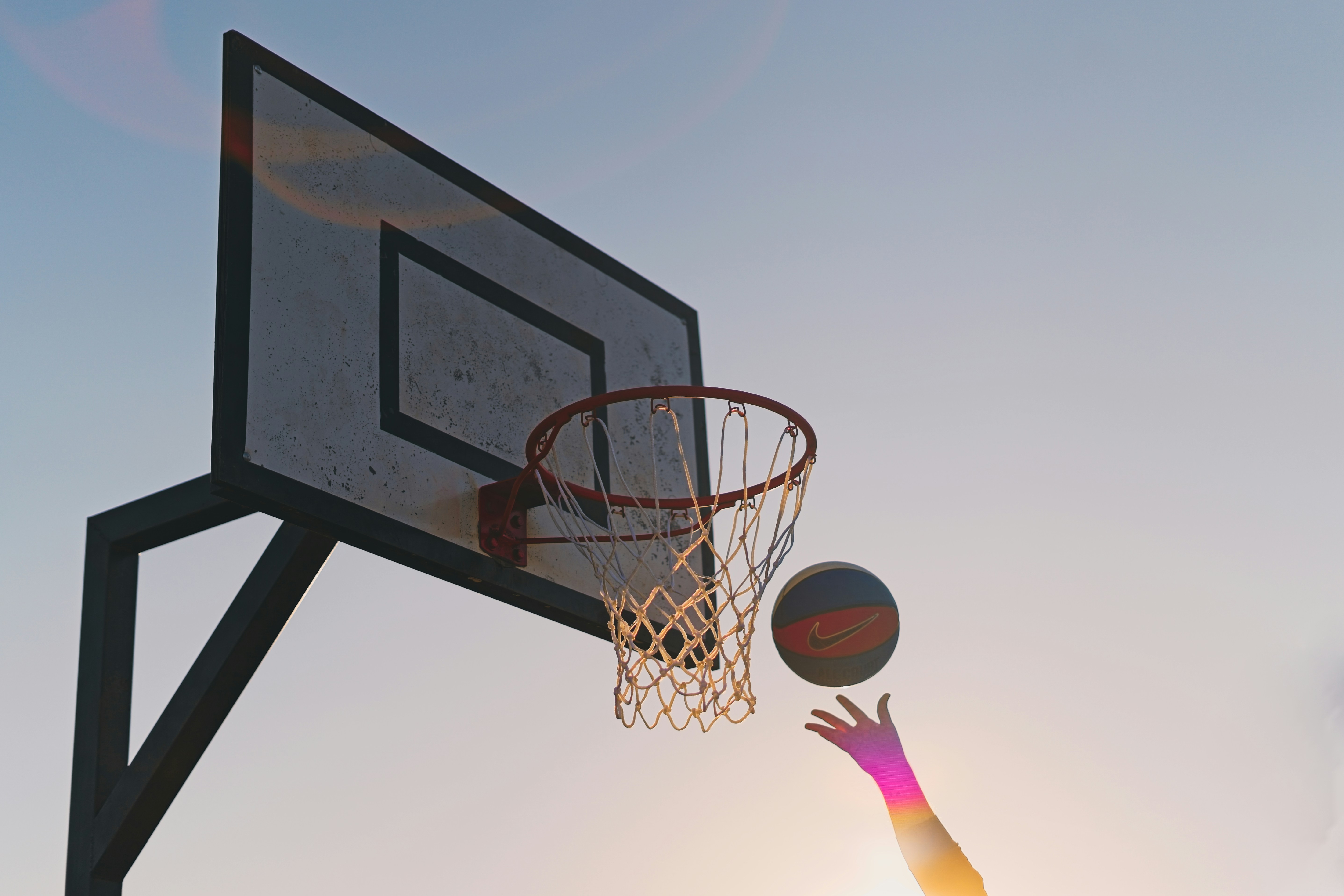 A basketball airborne near an outdoor hoop and net, guided by an outstretched hand bathed in warm sunset lens flare