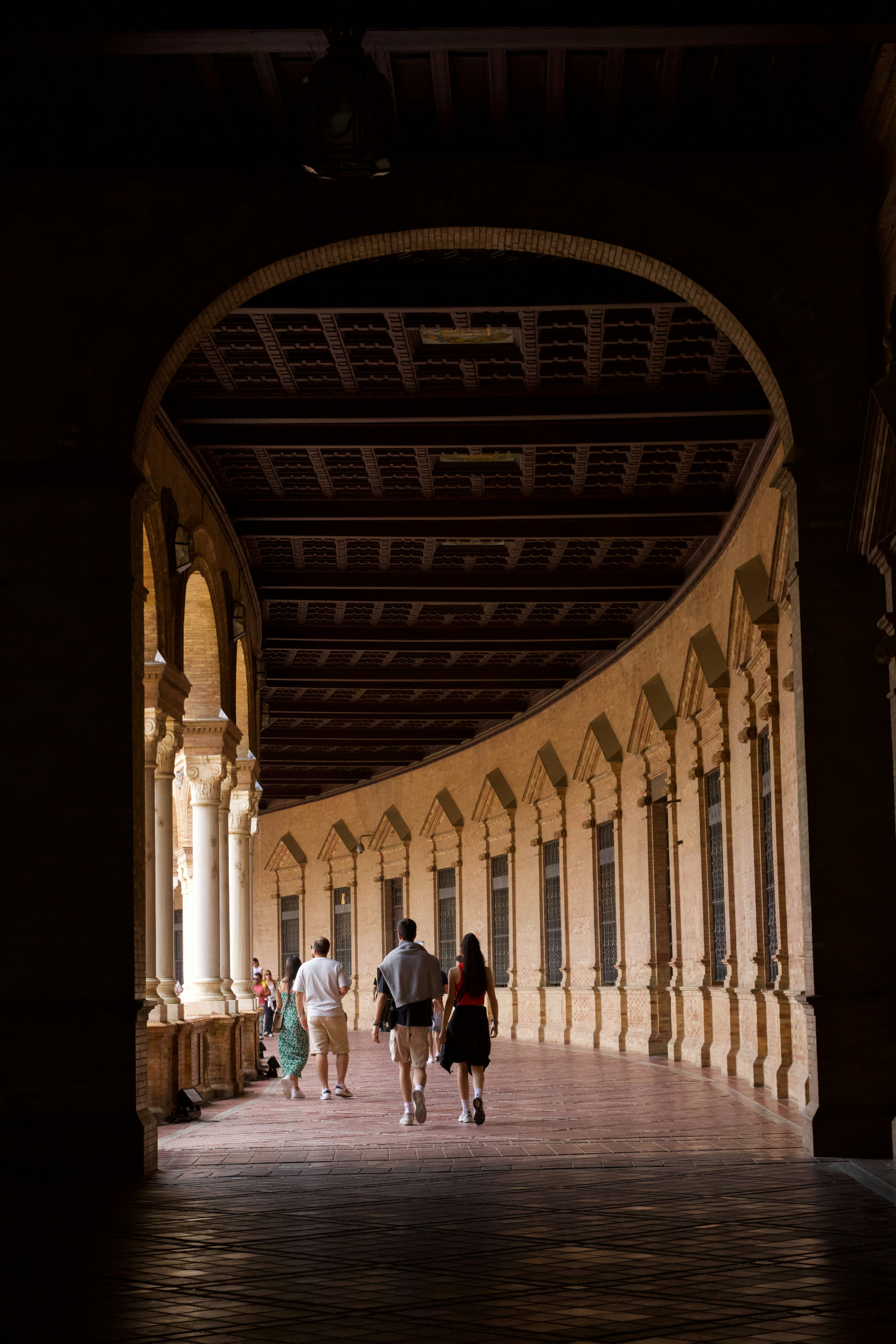 Sunlit figures walk along a curved arcade framed by repeating arches. The composition highlights warm stone architecture and a soft, morning glow along the corridor.