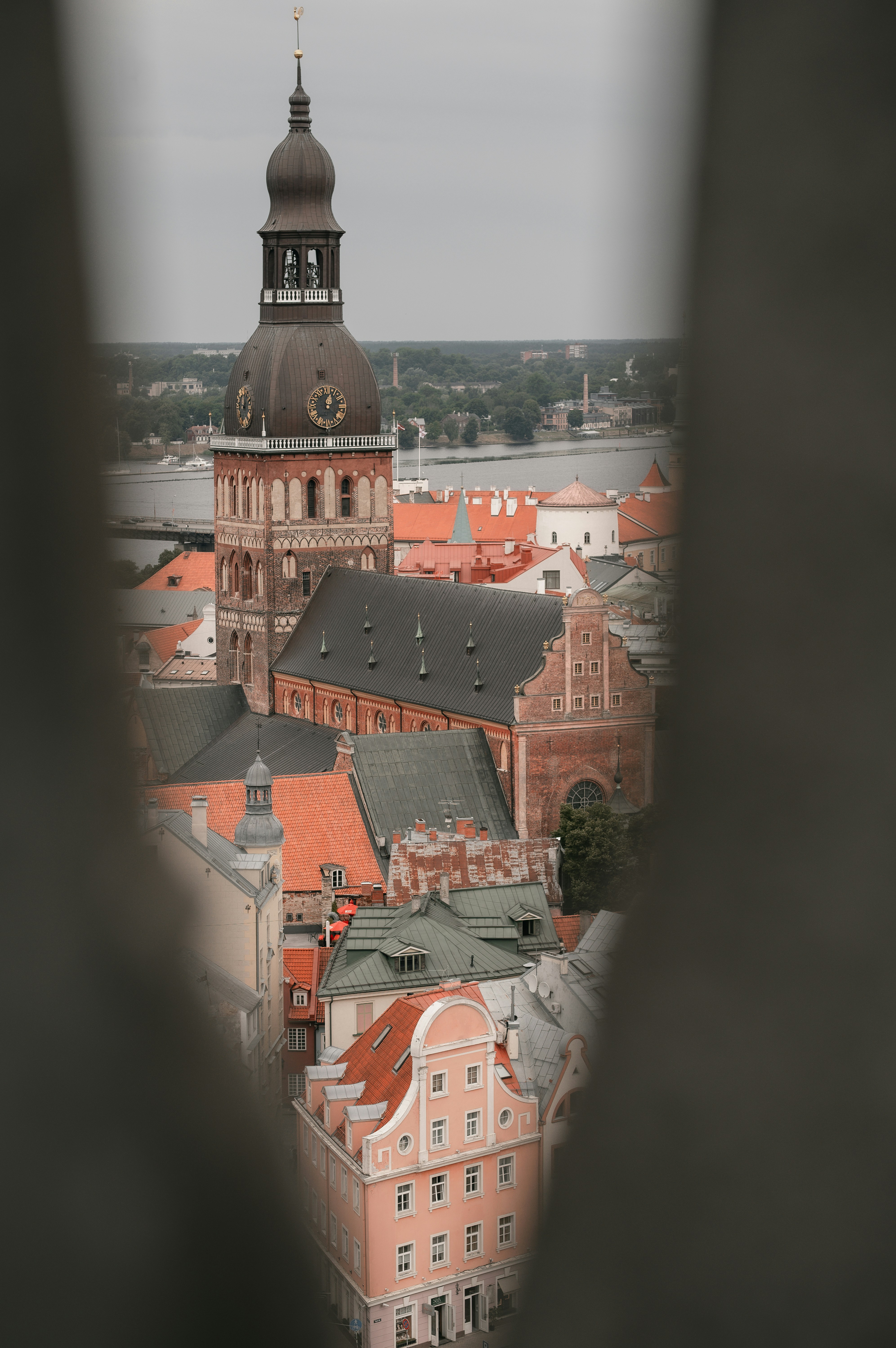Brick clock tower rises above red-tiled roofs, framed by blurred foreground arches and a distant river skyline.