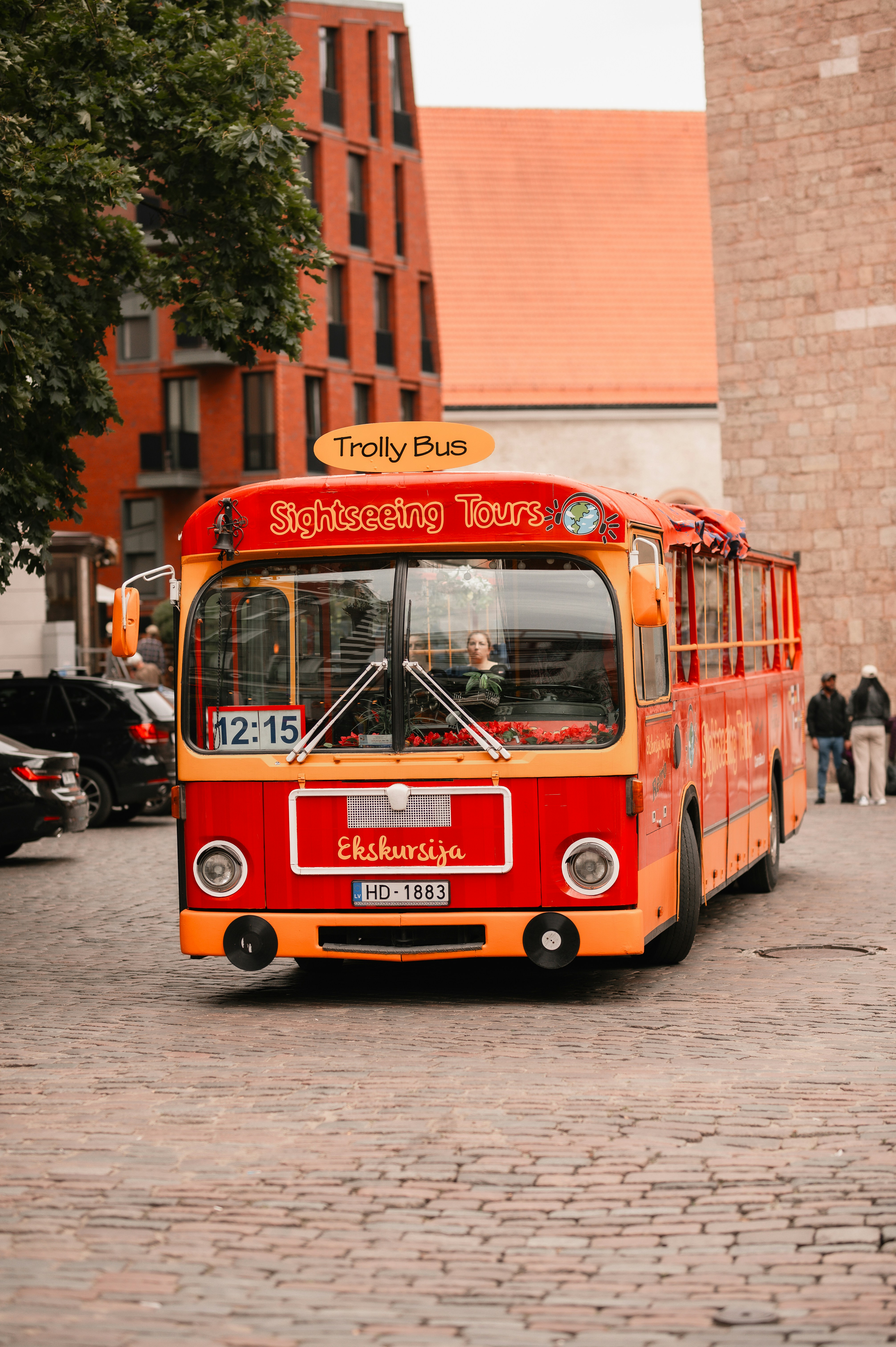 Bright orange sightseeing bus on a cobblestone street with Historic buildings in the background; passengers visible through the front windows. This is a photograph capturing a street scene with a traveling tour bus.