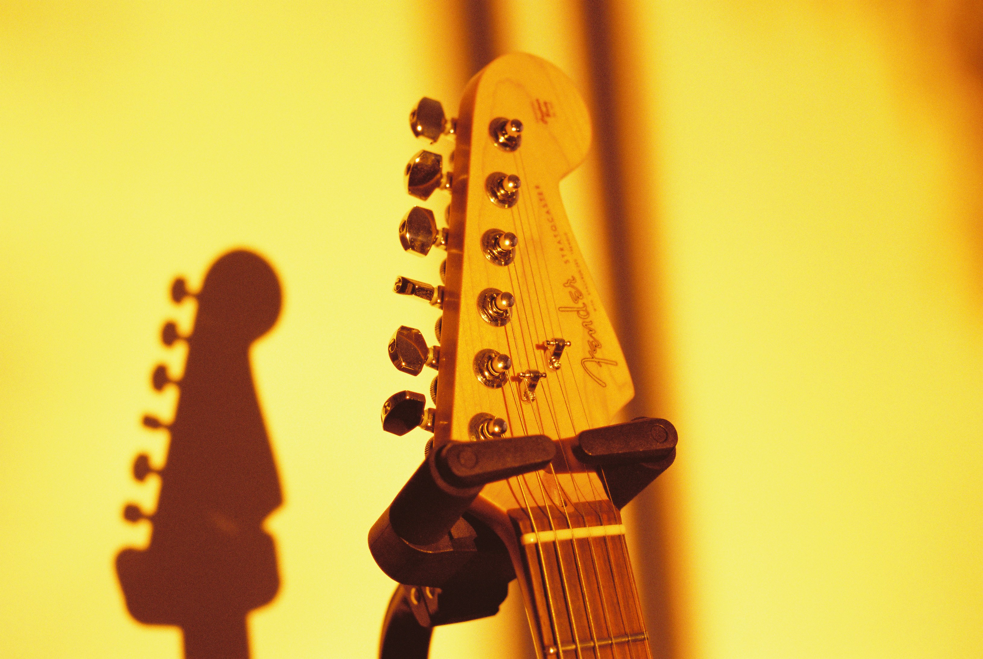 Warm-lit guitar headstock silhouette with a strong shadow on the left; the focus is the tuners and logo area against a yellow-orange background.